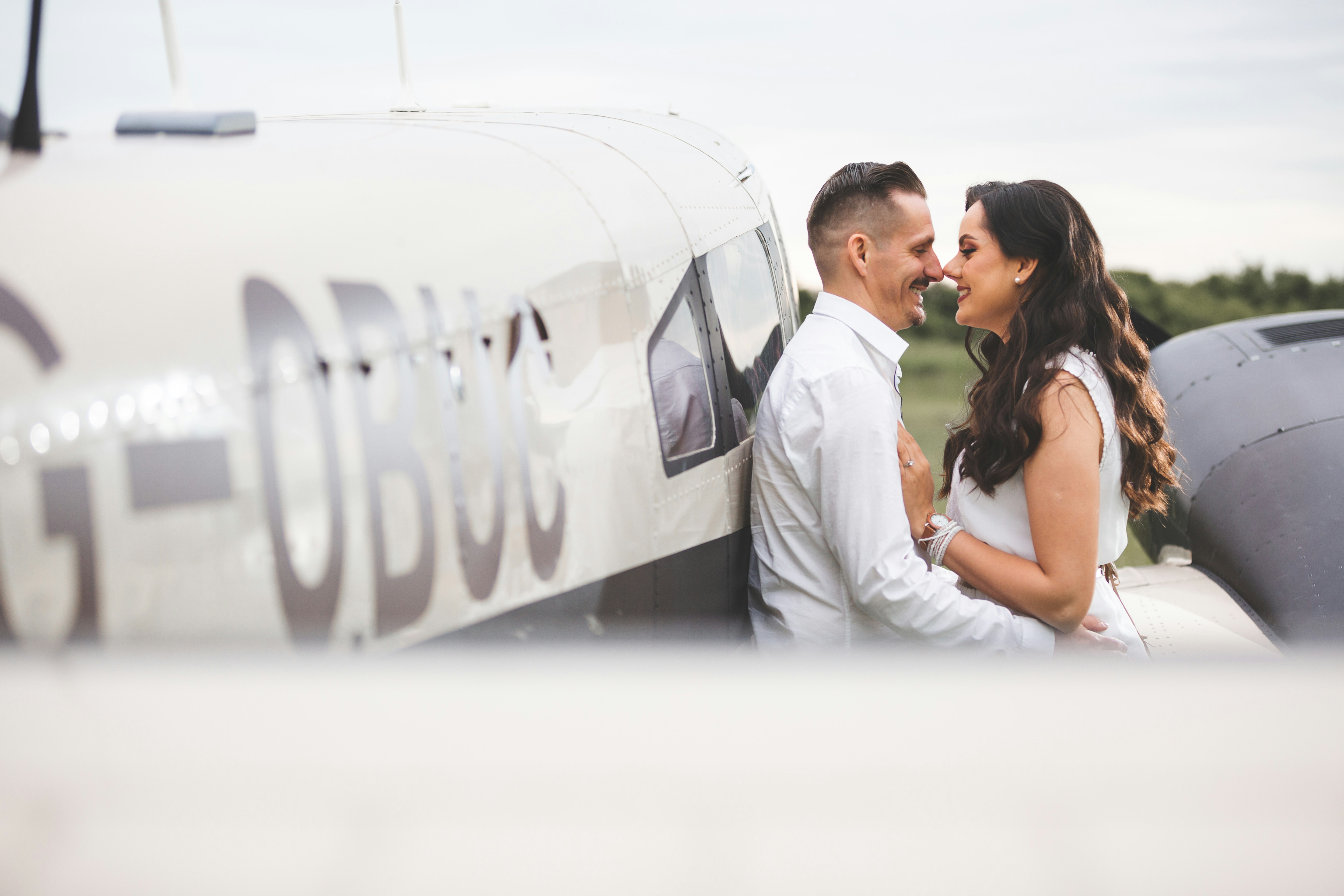 Woman and her fiance close together, nose to nose, next to a private plane, both laughing. | Couple embracing by a small airplane