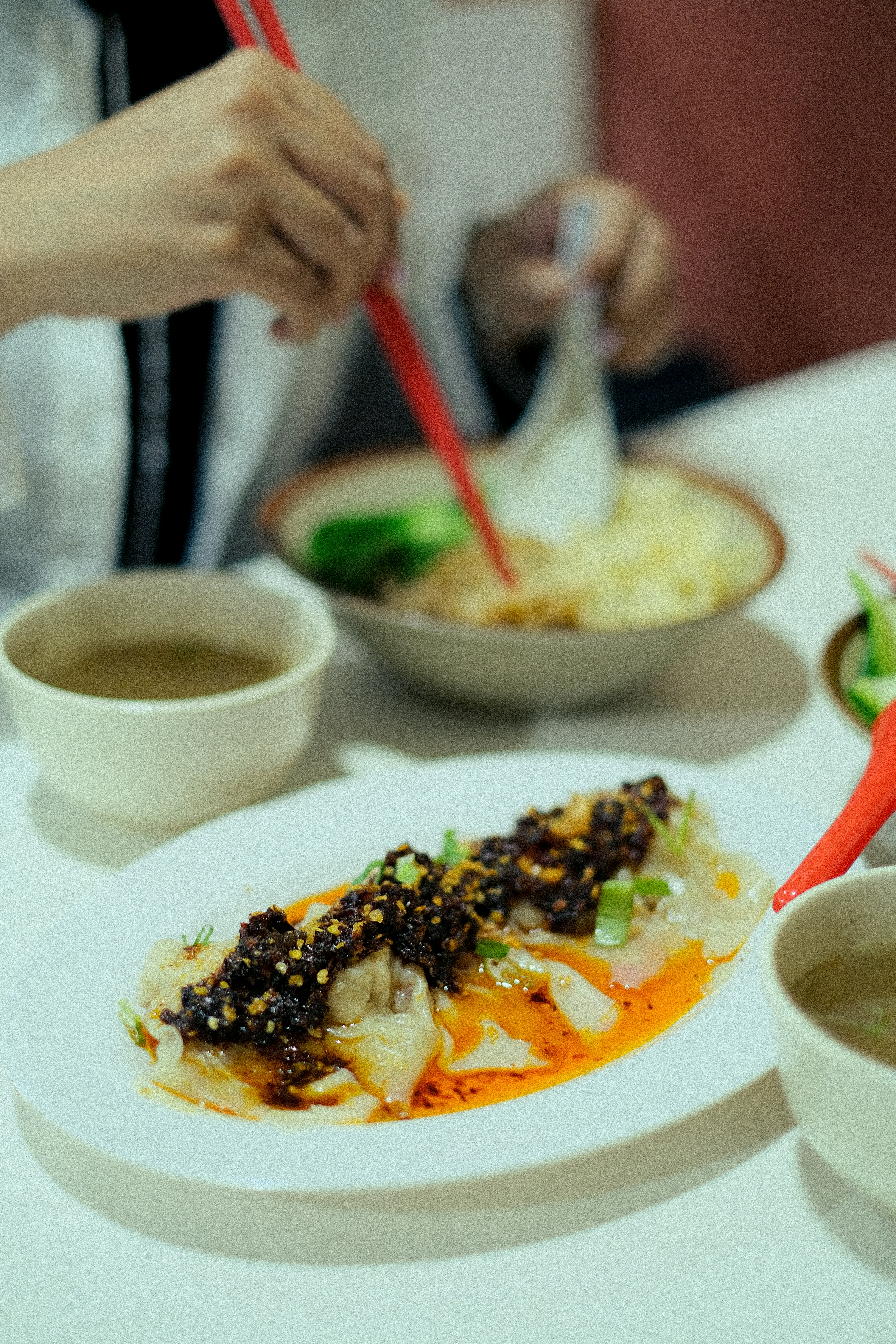 Delicious plate of dumplings drizzled with spicy sauce, accompanied by a bowl of noodles and broth. The scene captures the joy of dining with chopsticks in hand.
