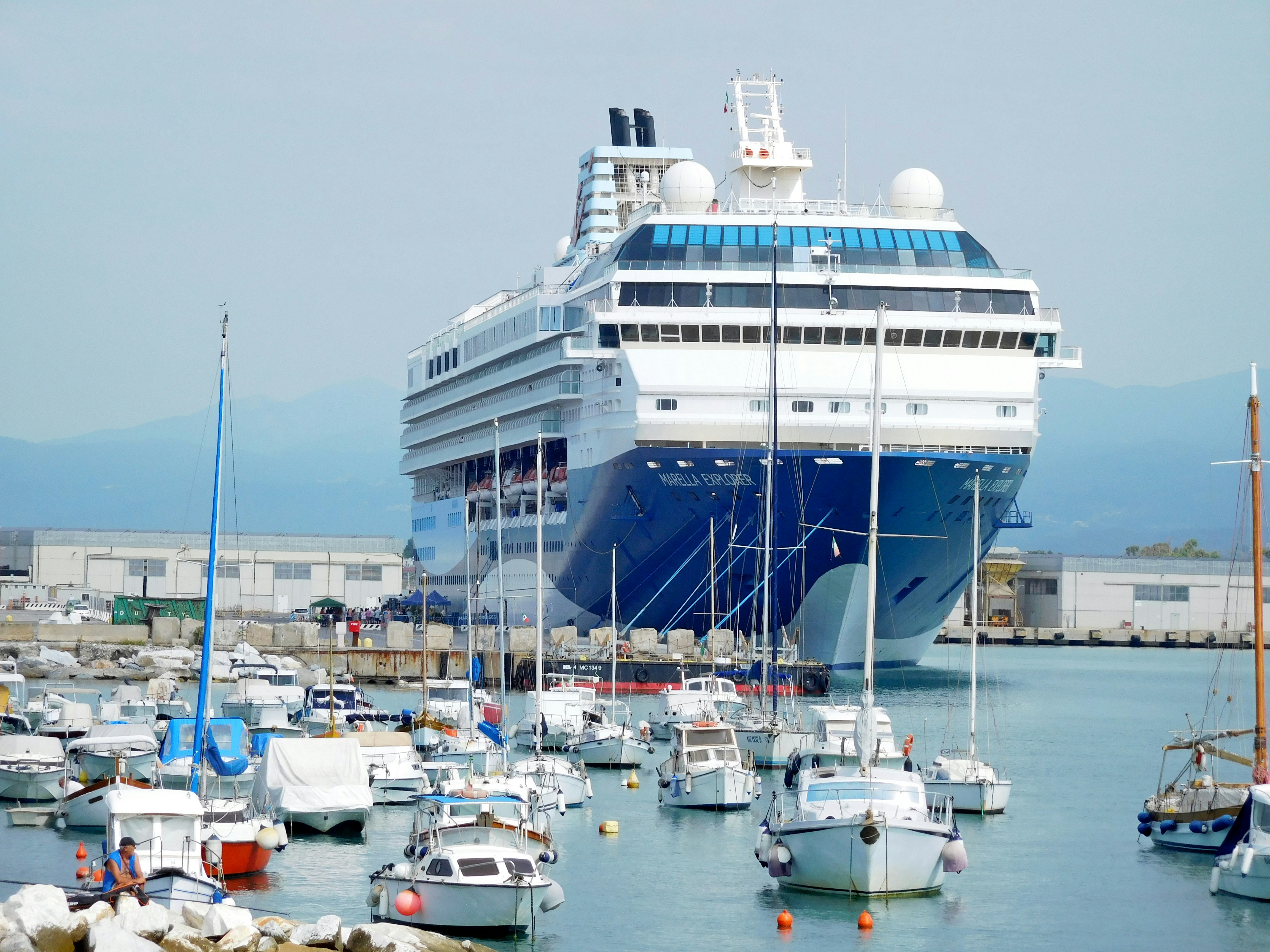 A large cruise ship docked in a busy harbor surrounded by smaller sailboats, showcasing a blend of maritime activity. The serene water reflects the vessels and the distant mountains.