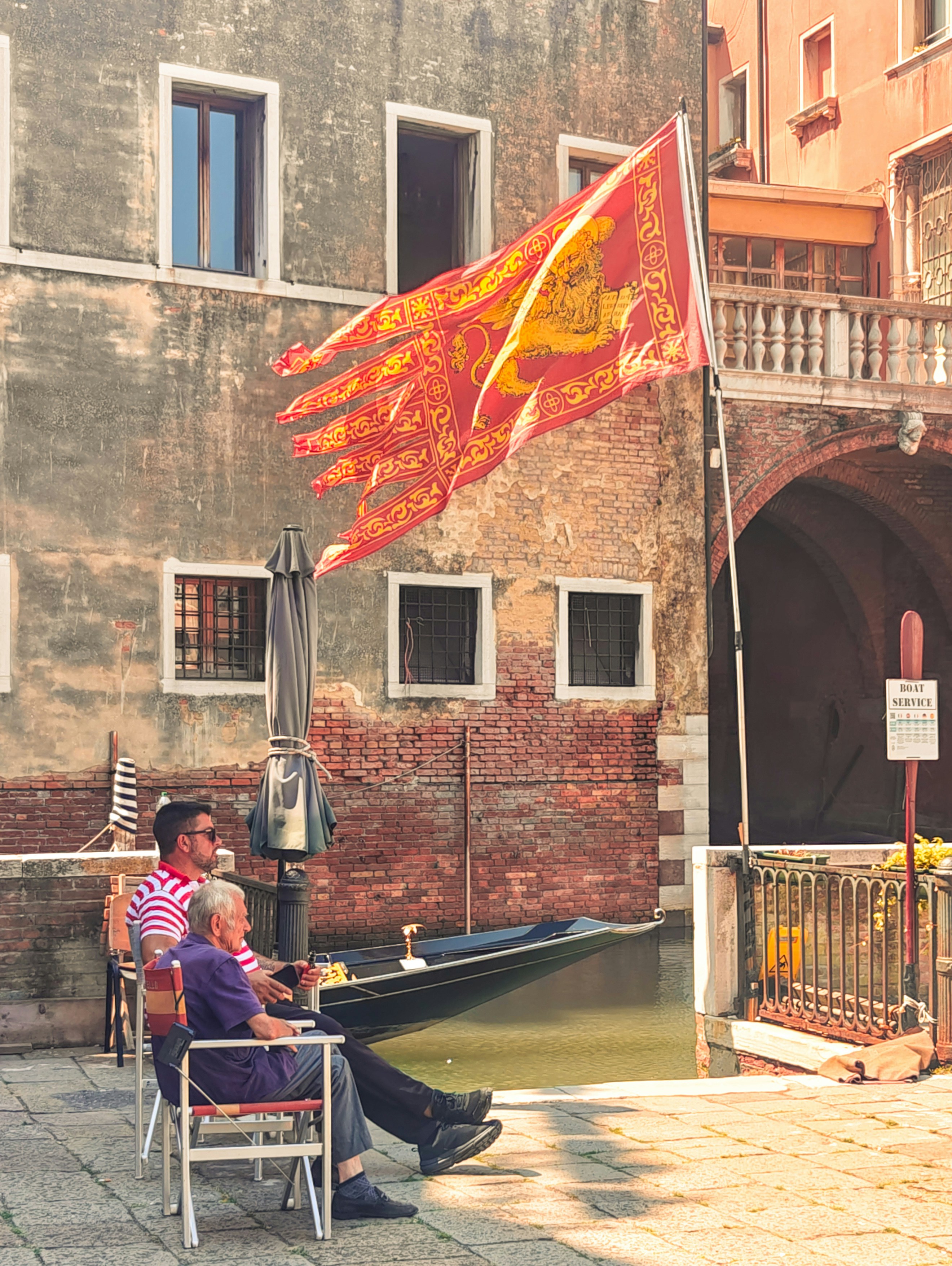 Two men relax by a canal under a venetian flag.