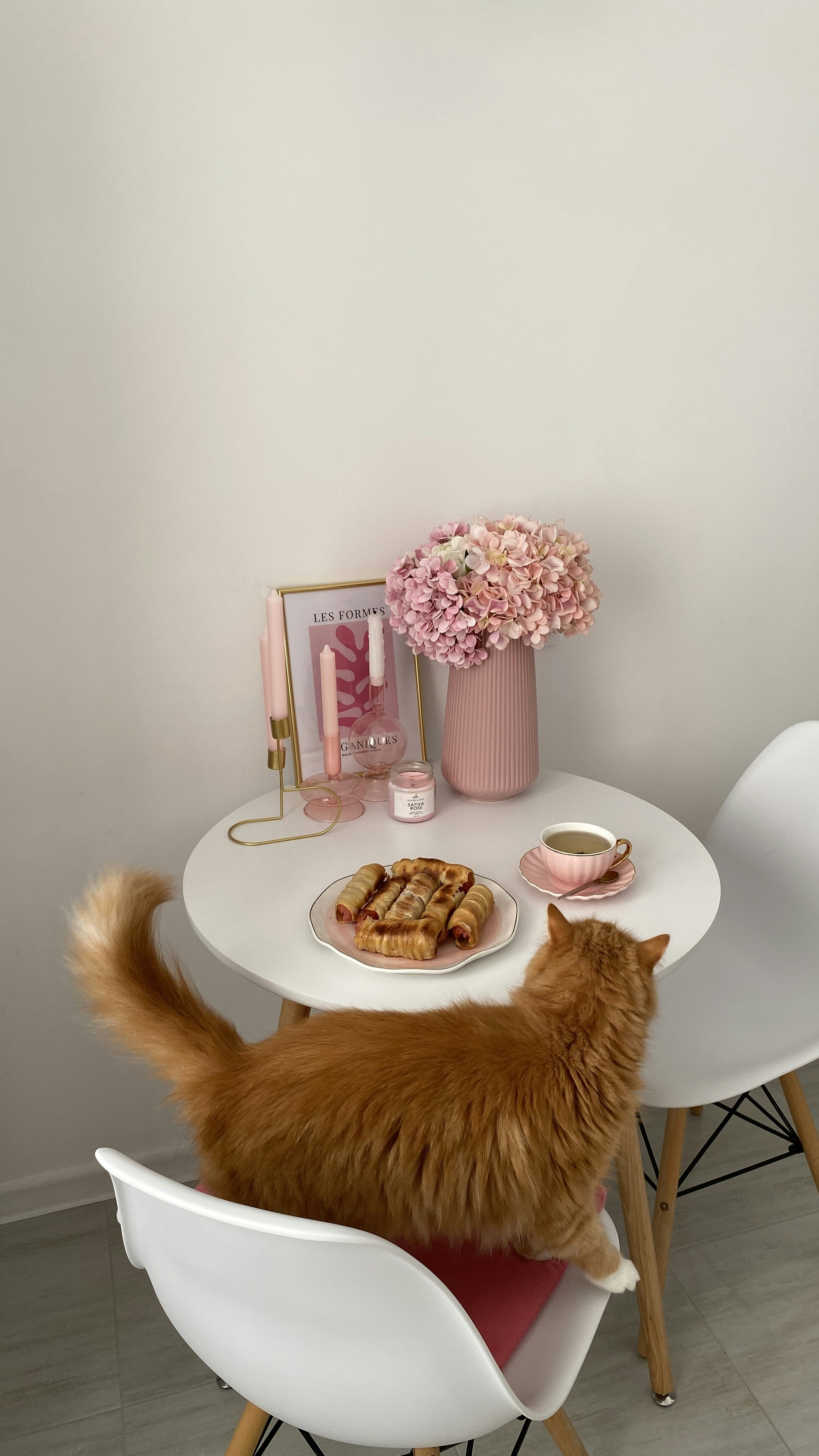 A fluffy orange cat explores a stylish pink-themed breakfast table adorned with pastries, a delicate cup, and a floral centerpiece.