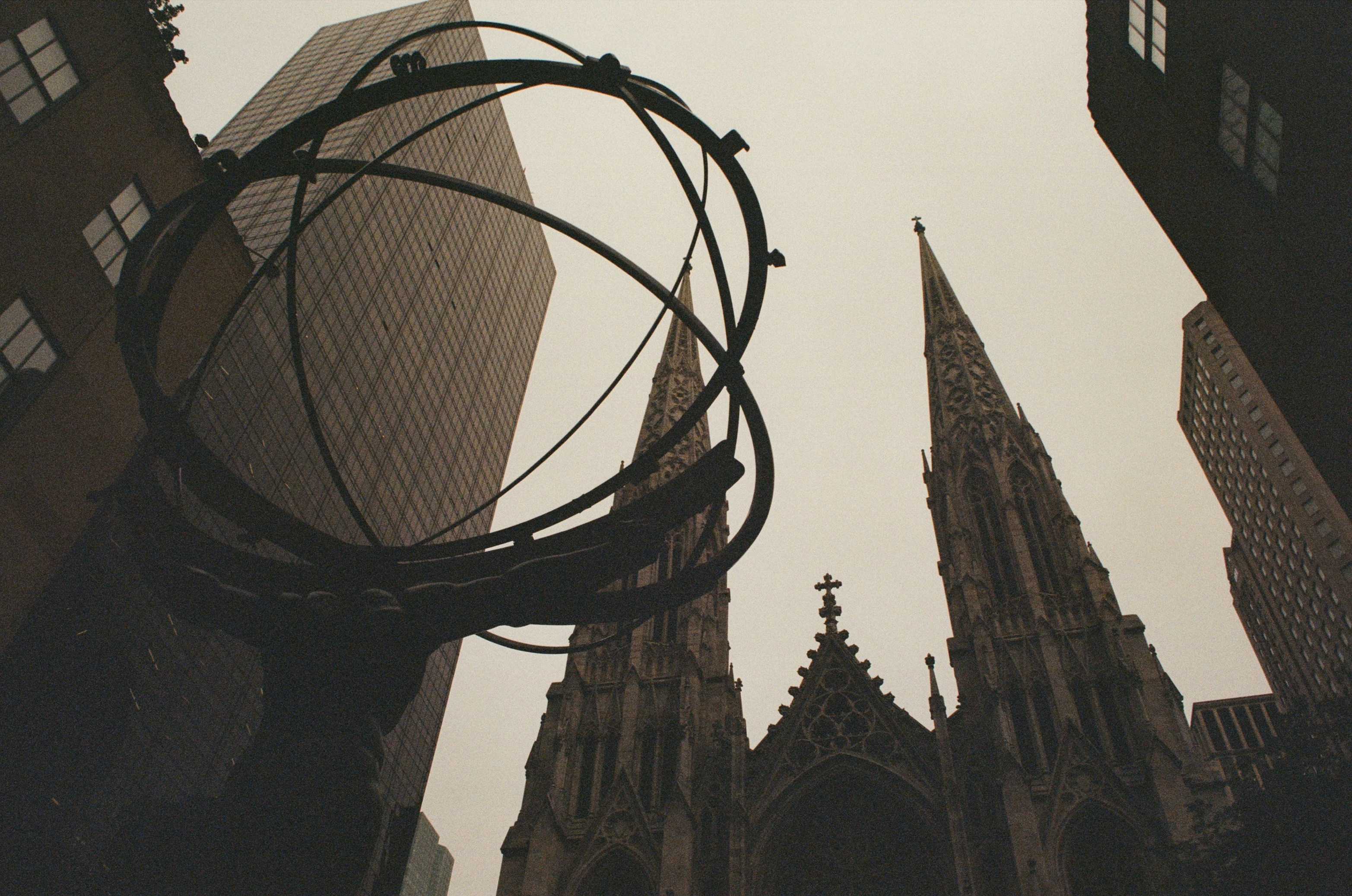 A dramatic low-angle view showcasing a globe sculpture framed by towering skyscrapers and the spires of a historic cathedral. The scene captures the contrast between modern and classic architecture.