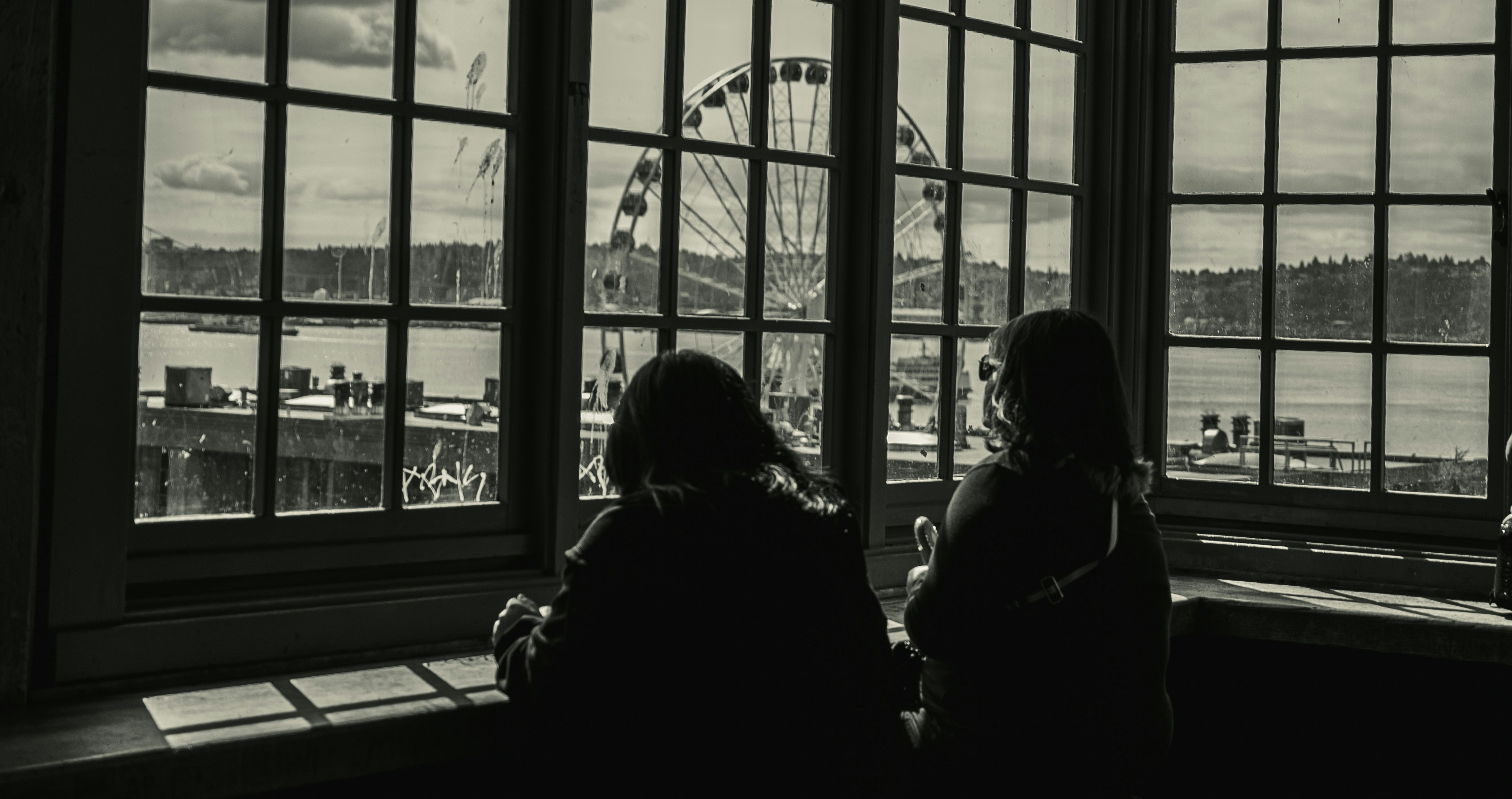 Two figures gazing out the window at a distant ferris wheel and waterfront, framed by a grid of window panes.