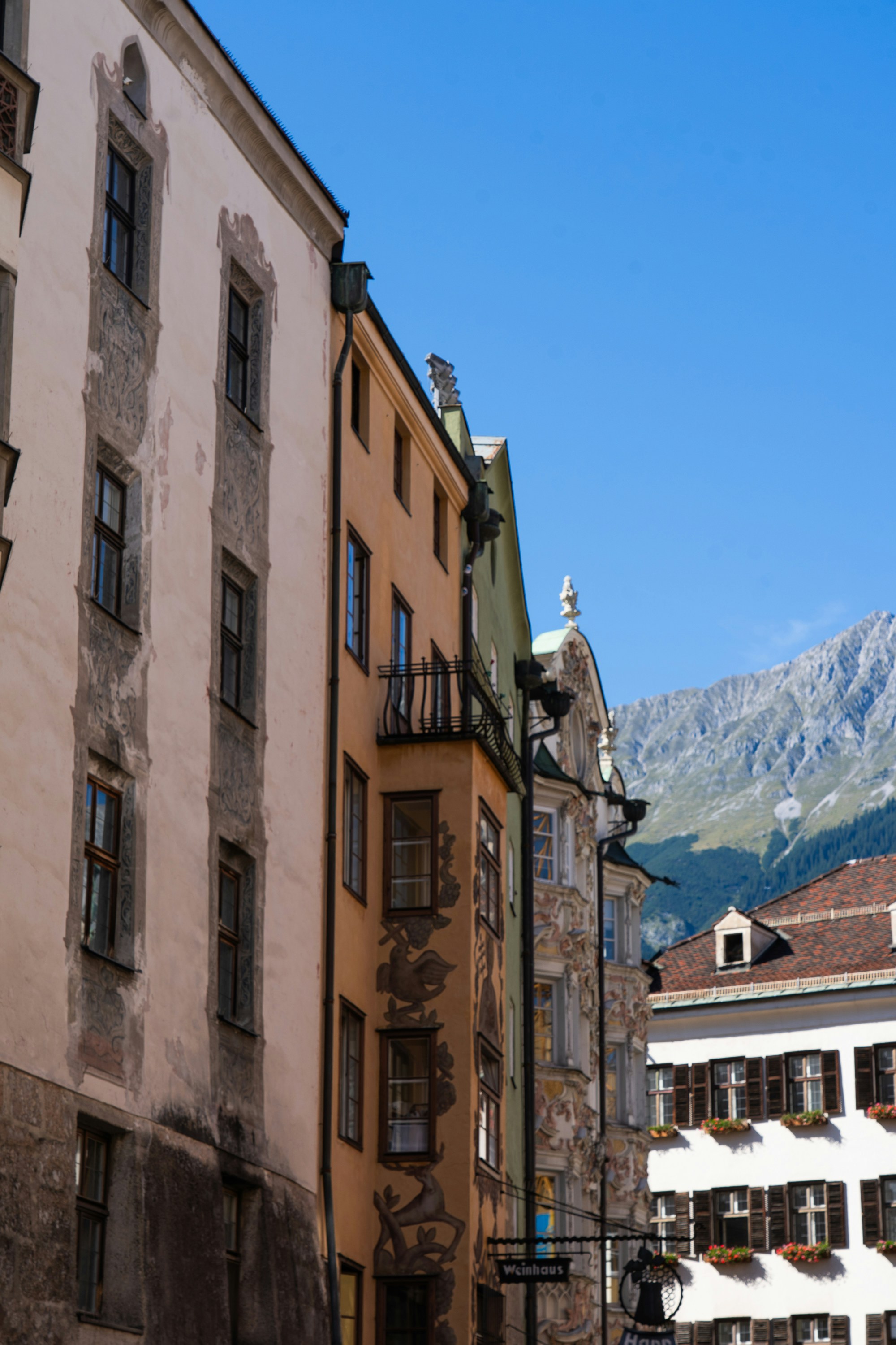 Charming buildings with intricate facades line a narrow street, framed by majestic mountains under a clear blue sky.