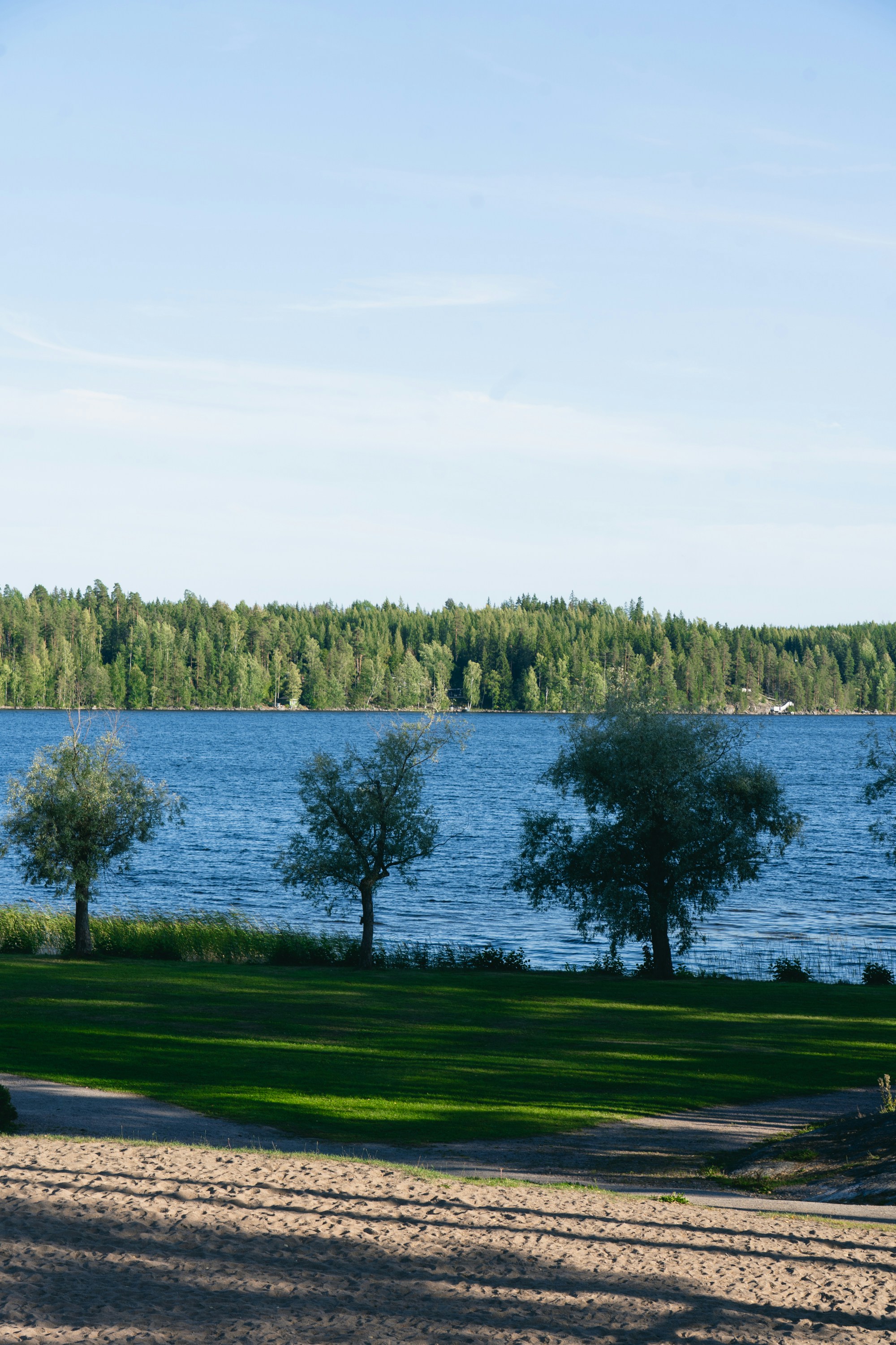 Lush green trees frame a calm lake under a clear blue sky, inviting relaxation and reflection. The sandy foreground adds a touch of warmth to the serene landscape.
