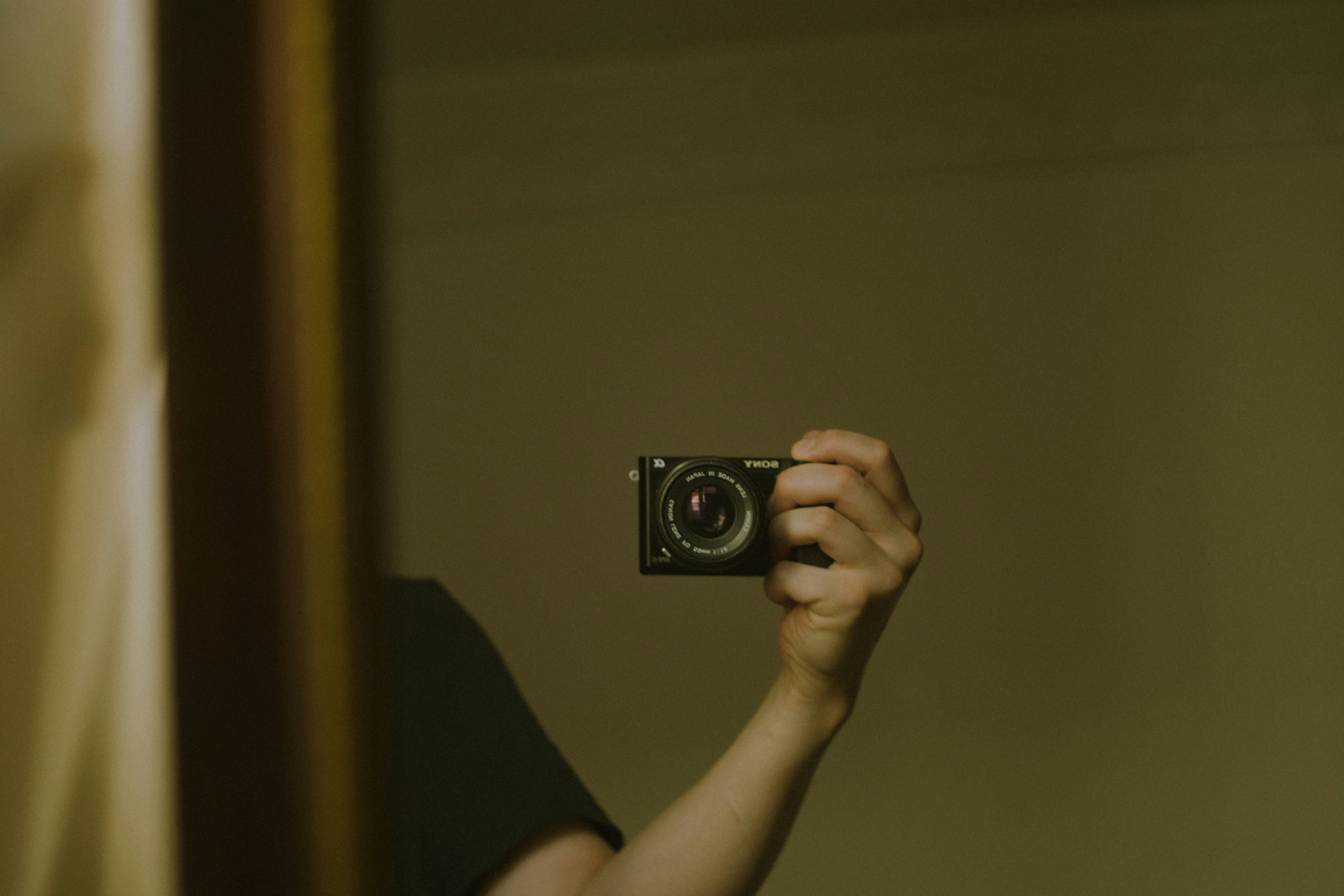 A hand holding a compact camera is reflected in a mirror, showcasing the interplay of light and shadow in a minimalist setting.