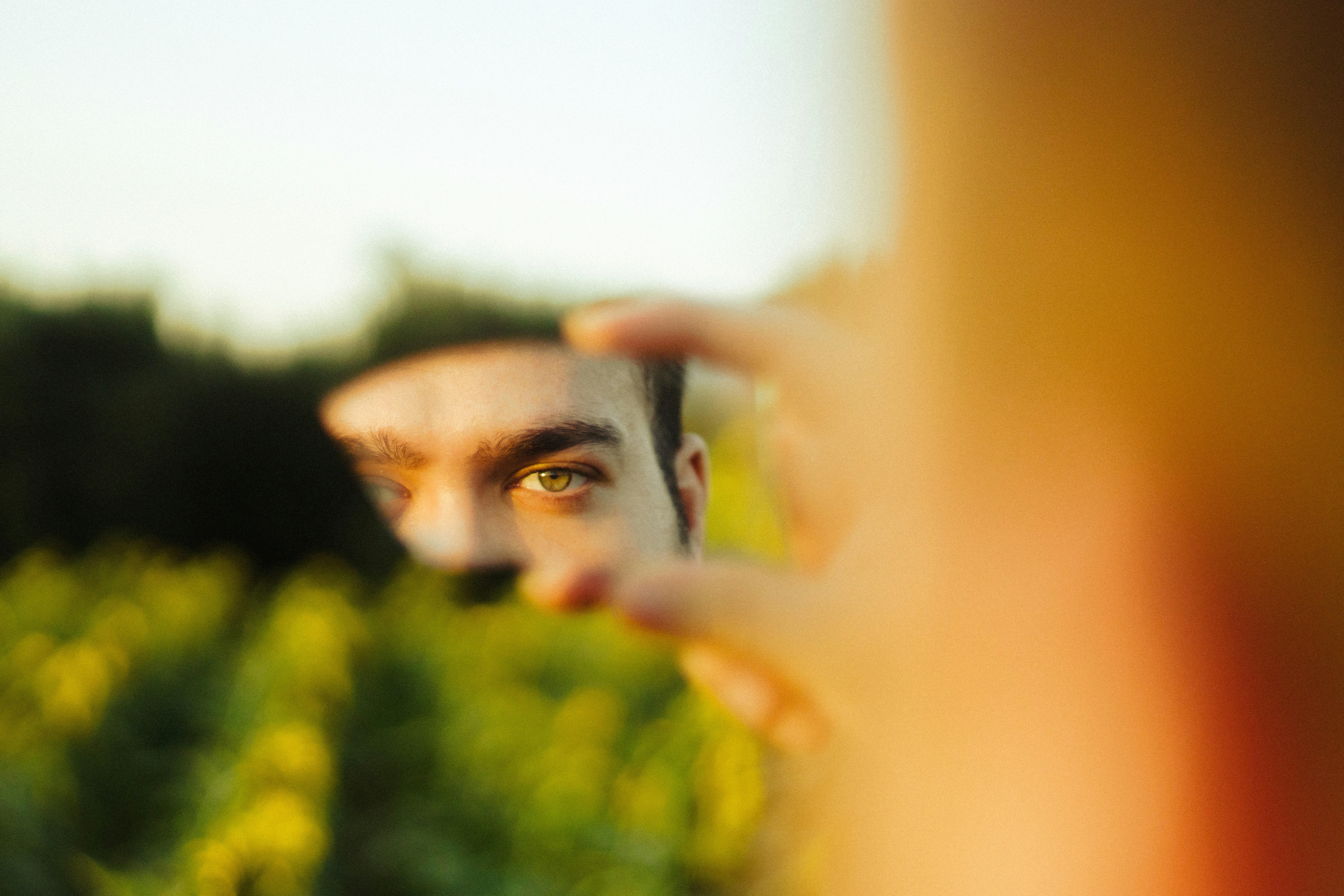A close-up of a person's eye reflected in a small mirror, surrounded by a blurred background of green foliage. The focus on the eye emphasizes introspection.