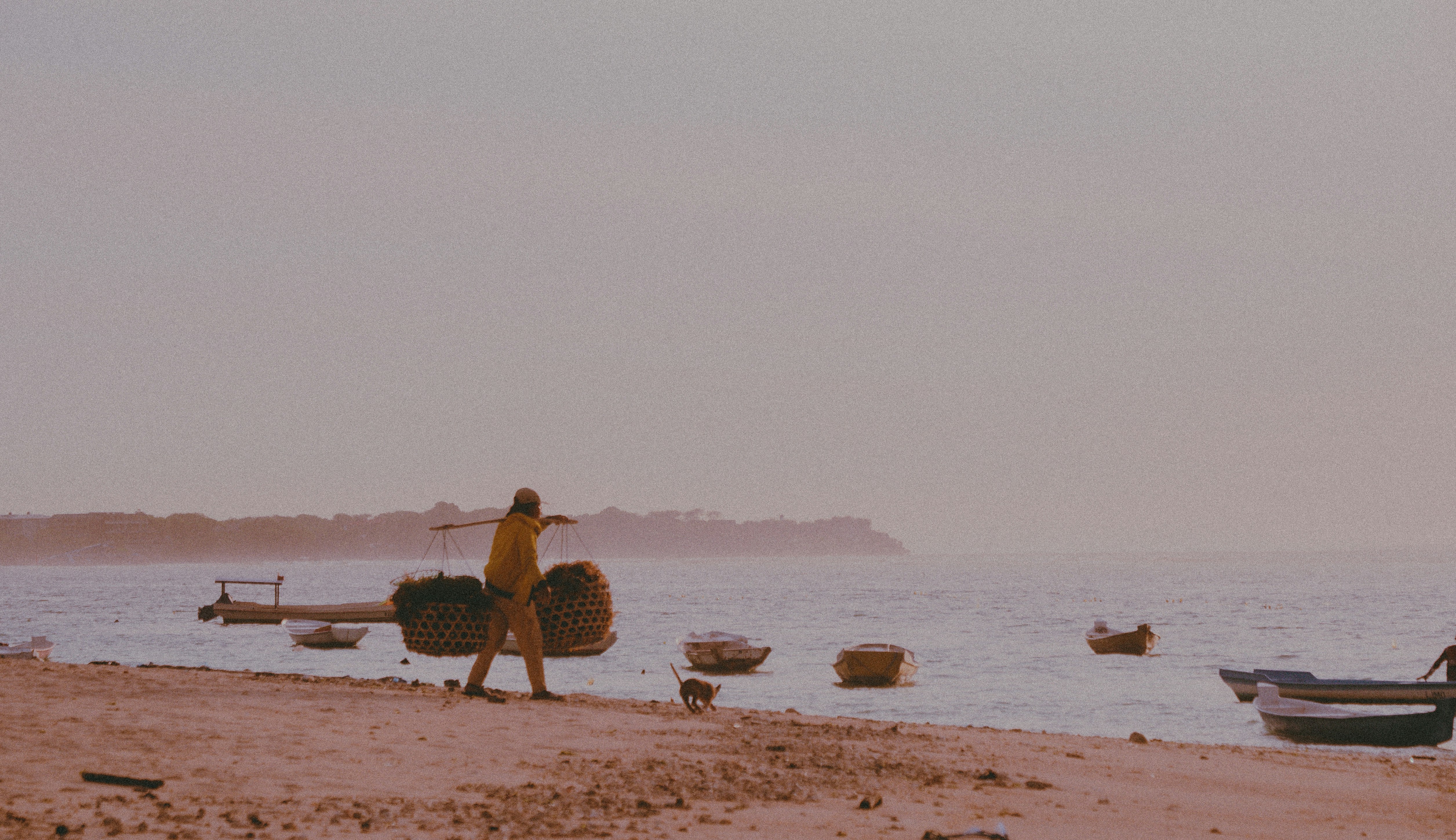 A person in a yellow coat carries baskets along a tranquil beach, with fishing boats gently bobbing on the water's surface.