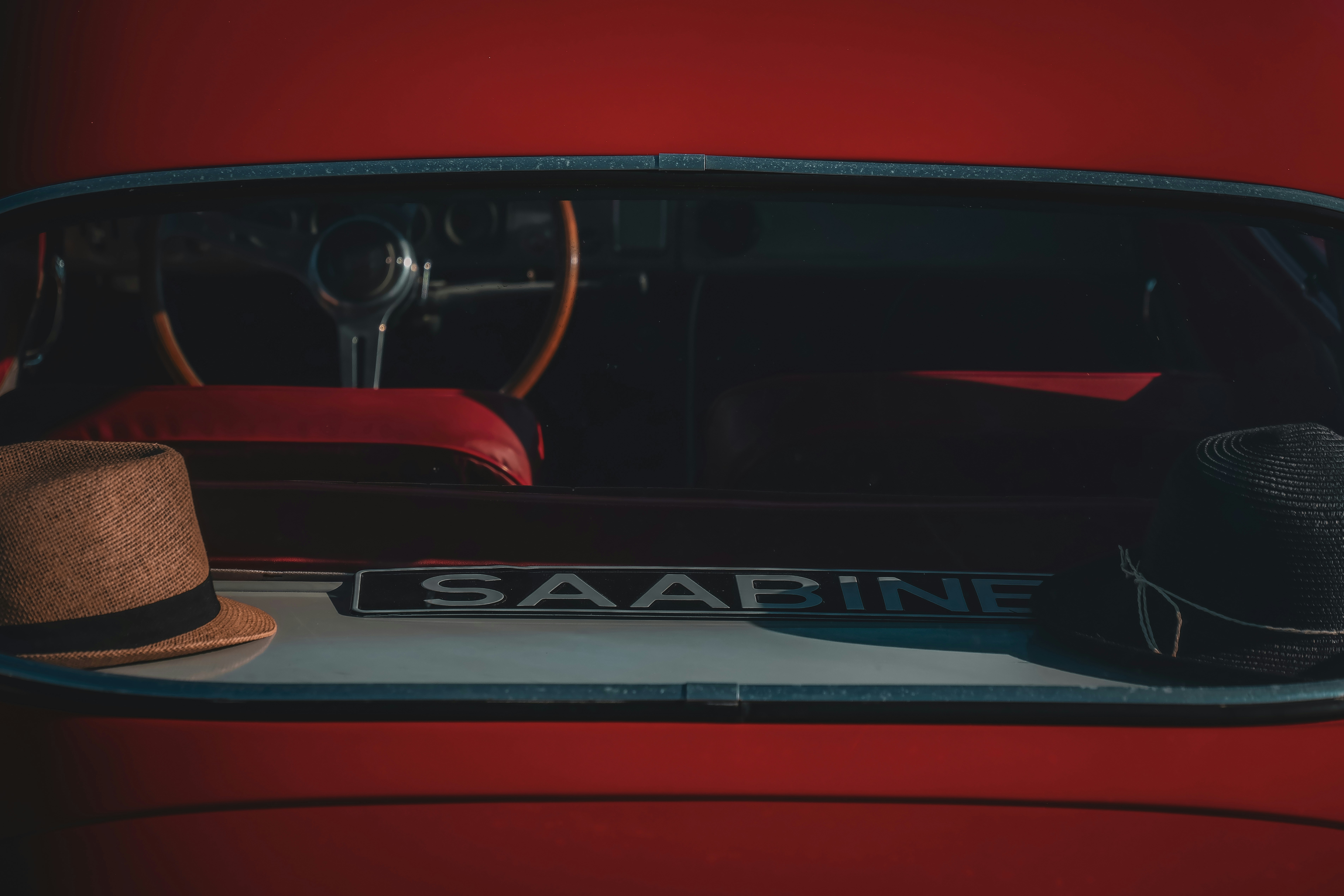 Through the rear window of a red vintage car, two classic fedoras rest side by side on the back shelf, framed by red leather seats and a wooden steering wheel. | Two hats on the trunk of a red car