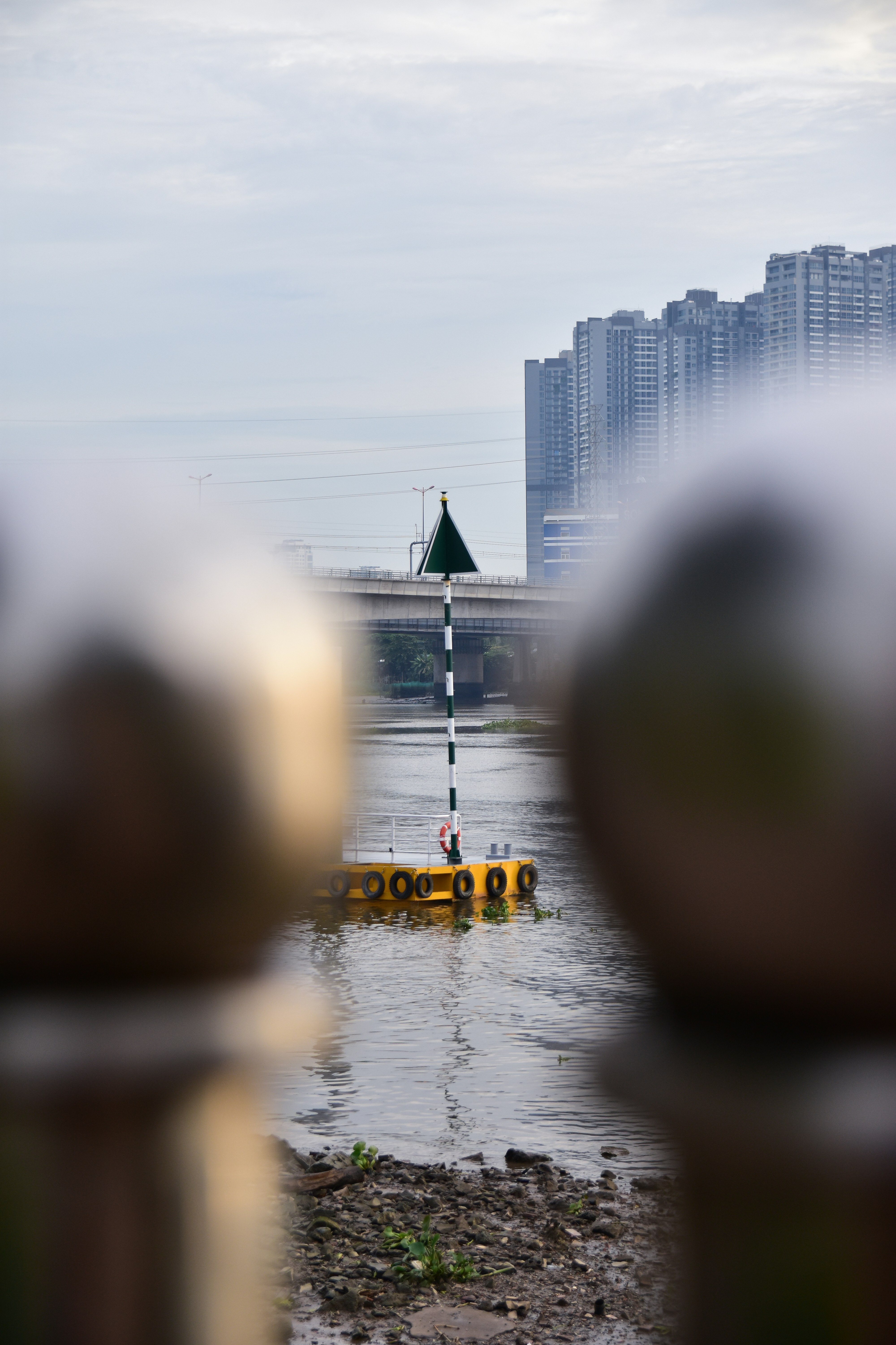 A brightly colored boat with a green triangular flag floats on a river, framed by blurred metallic posts, with urban buildings rising in the background.