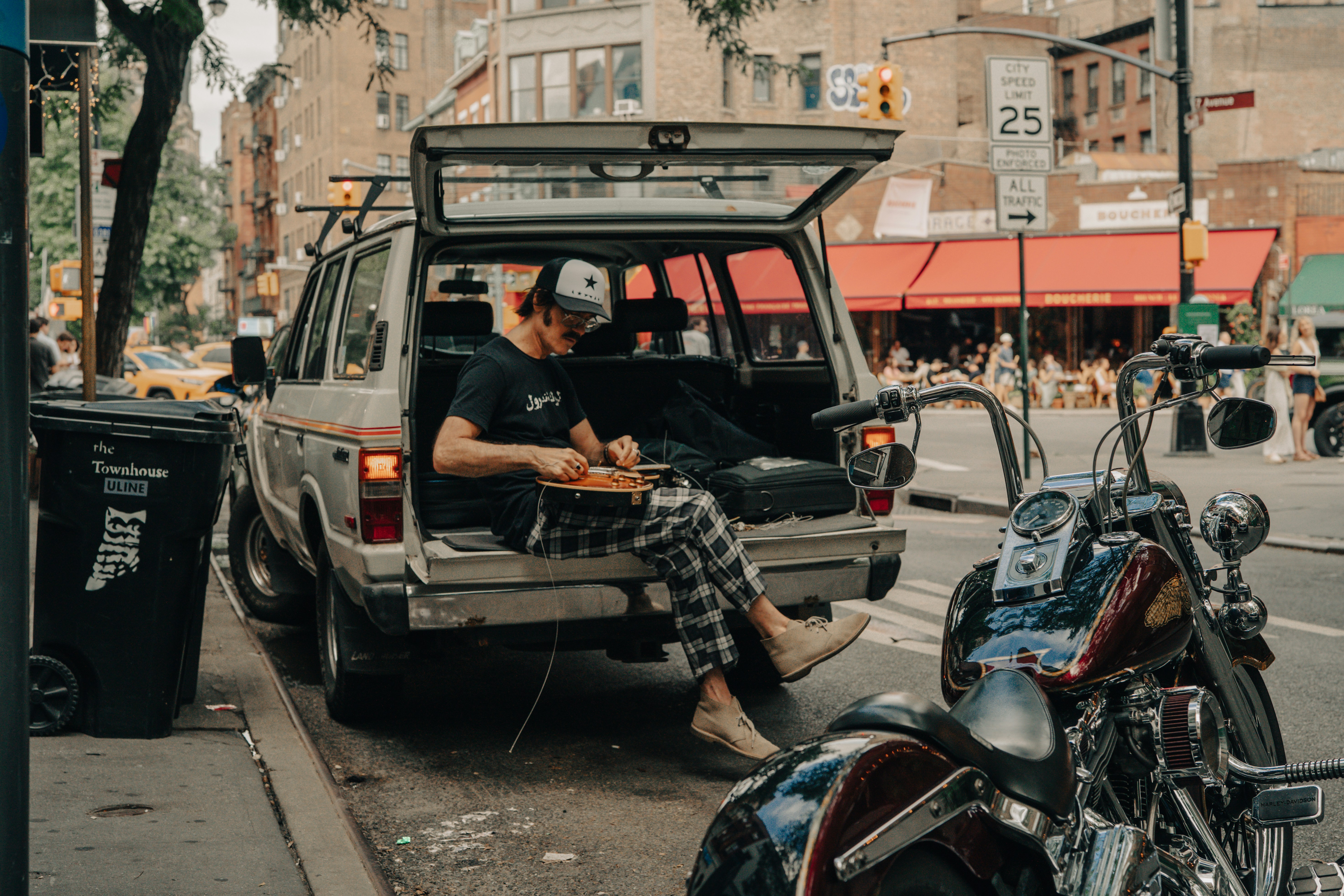 Man sitting on the tailgate of his car, re-stringing a guitar | Man sitting in open car trunk on city street.