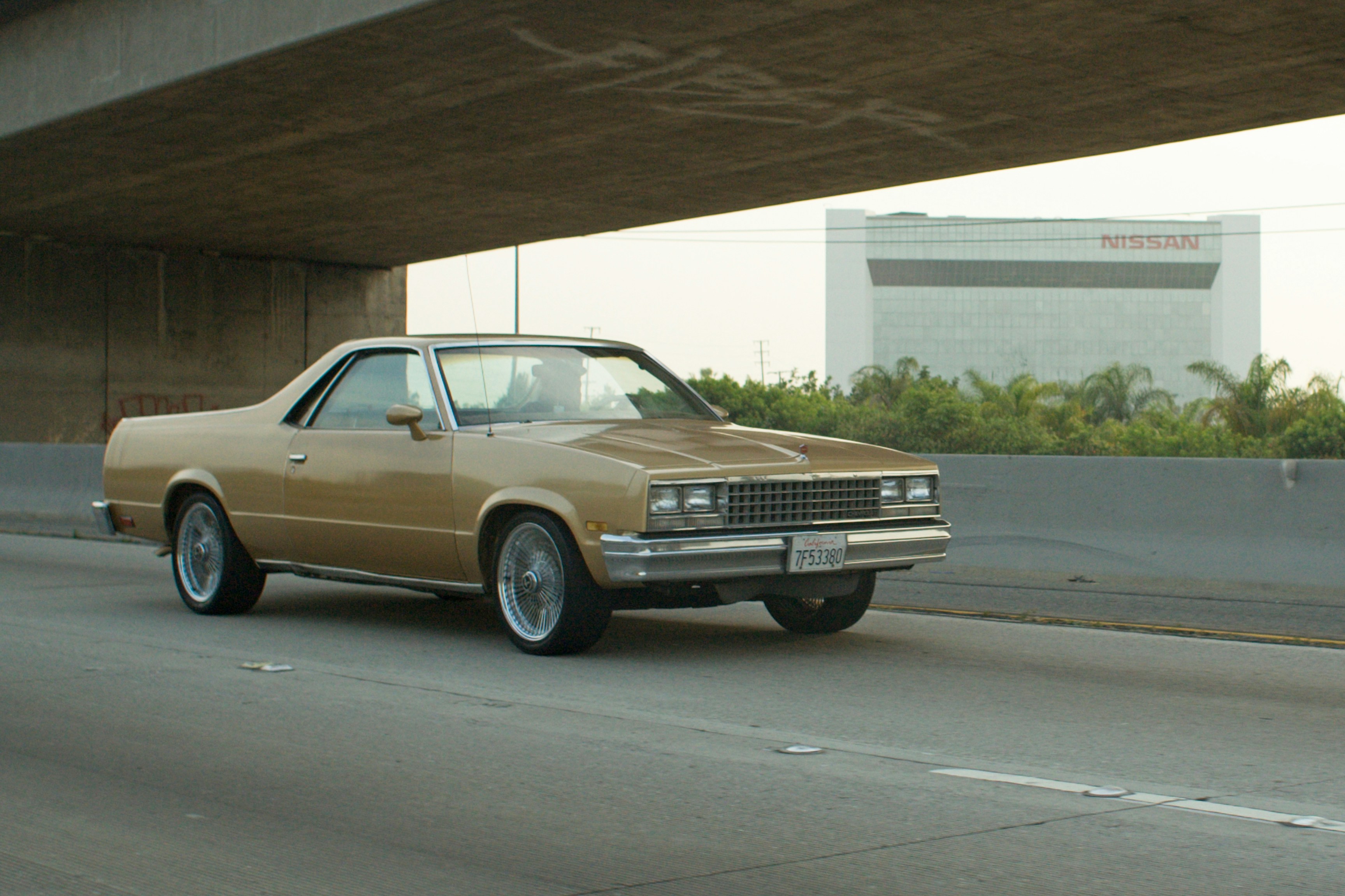 Classic gold El Camino cruising along the highway with industrial backdrop. The scene captures a blend of vintage style and modernity.