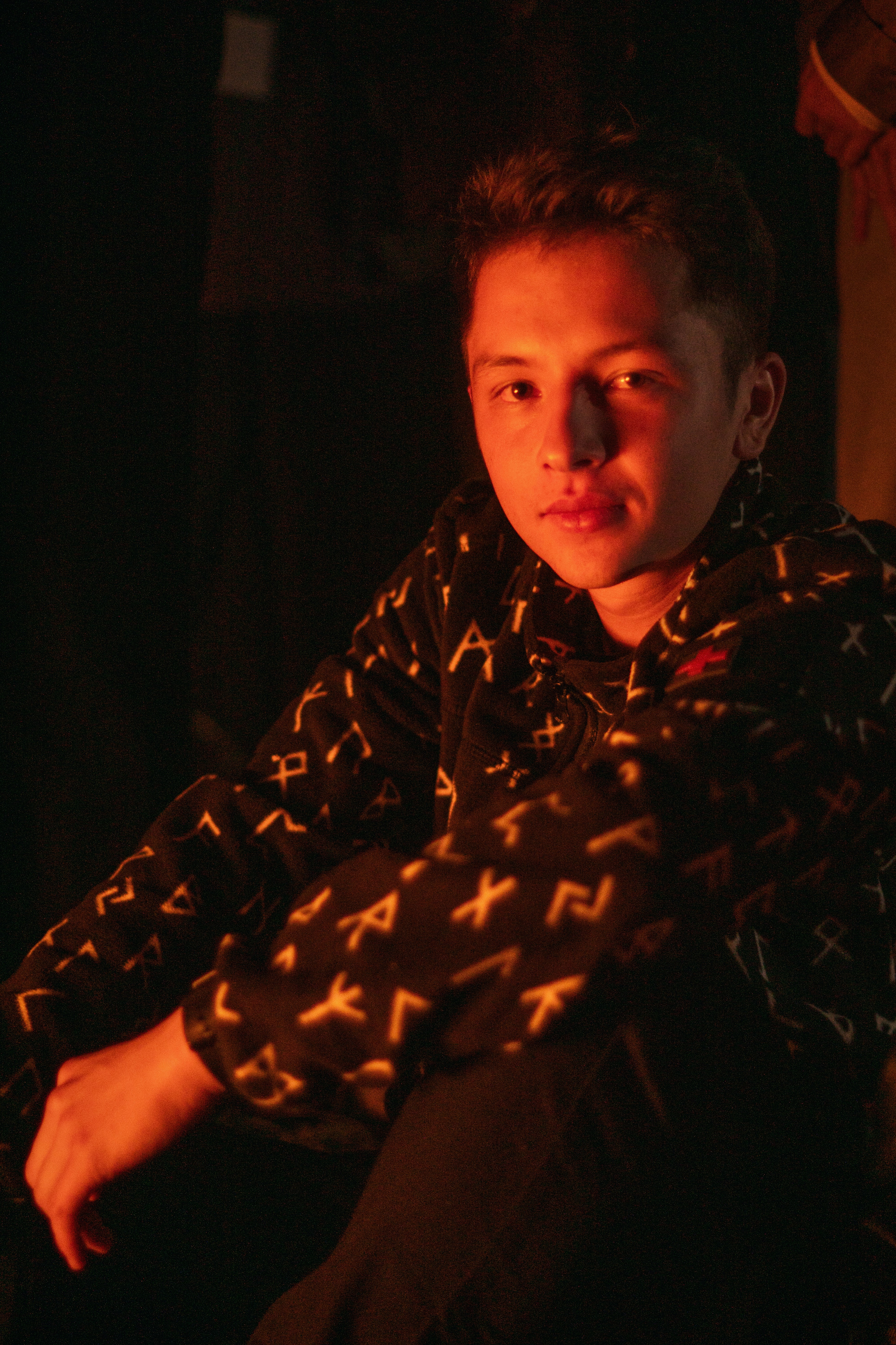 A man in a jacket is lit by the campfire light. | Young man in patterned shirt illuminated by firelight