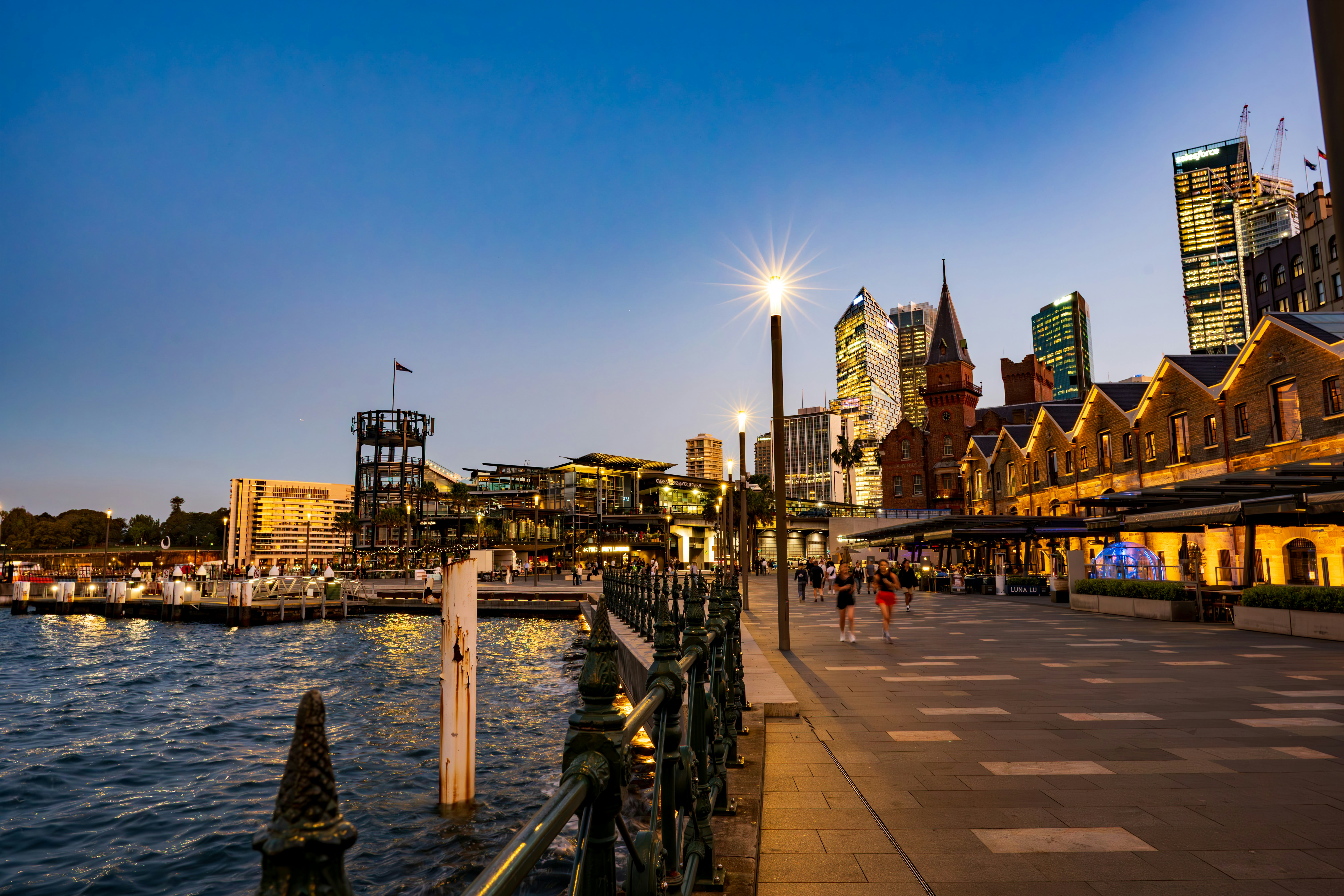 Vibrant waterfront scene at dusk, showcasing modern architecture alongside historic buildings, with soft light illuminating the promenade and water.