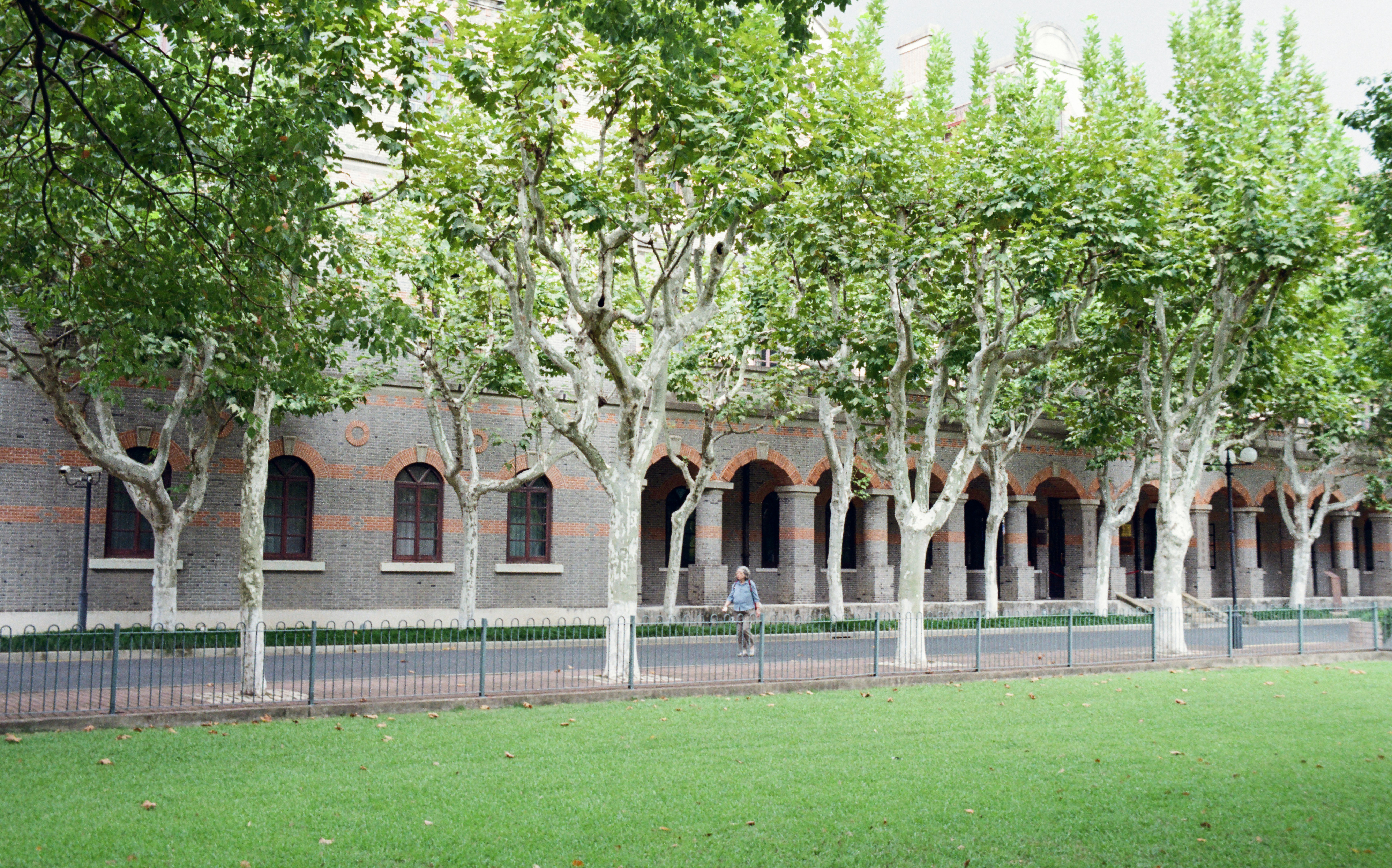 A person walking along a tree-lined pathway beside a historic brick building, showcasing architectural details and lush greenery.