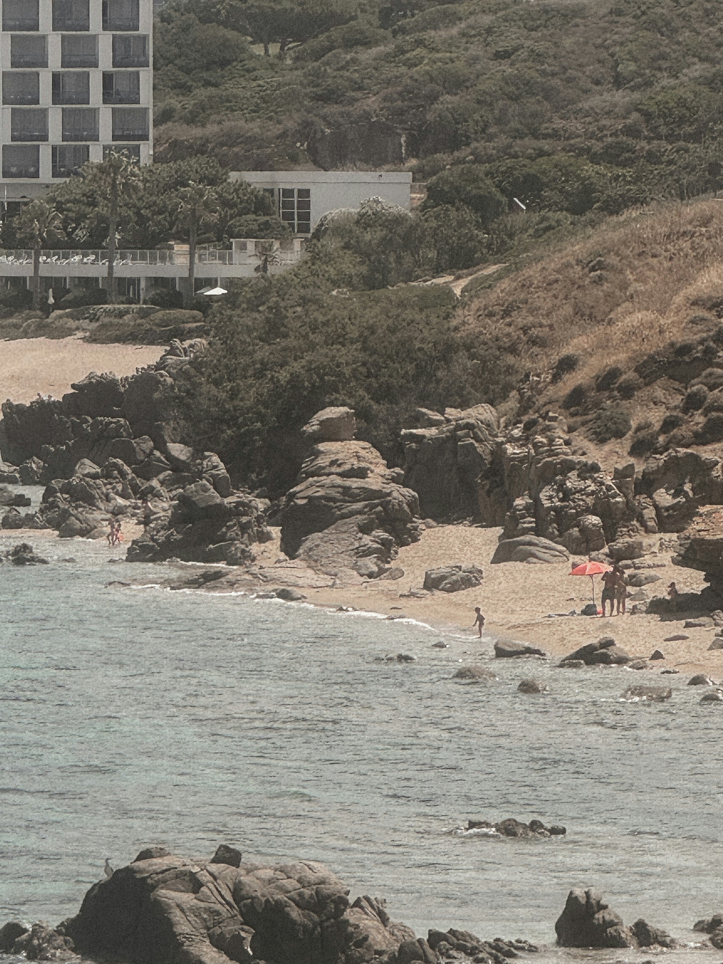 Plage parasol famille | Rocky beach with distant buildings and trees.