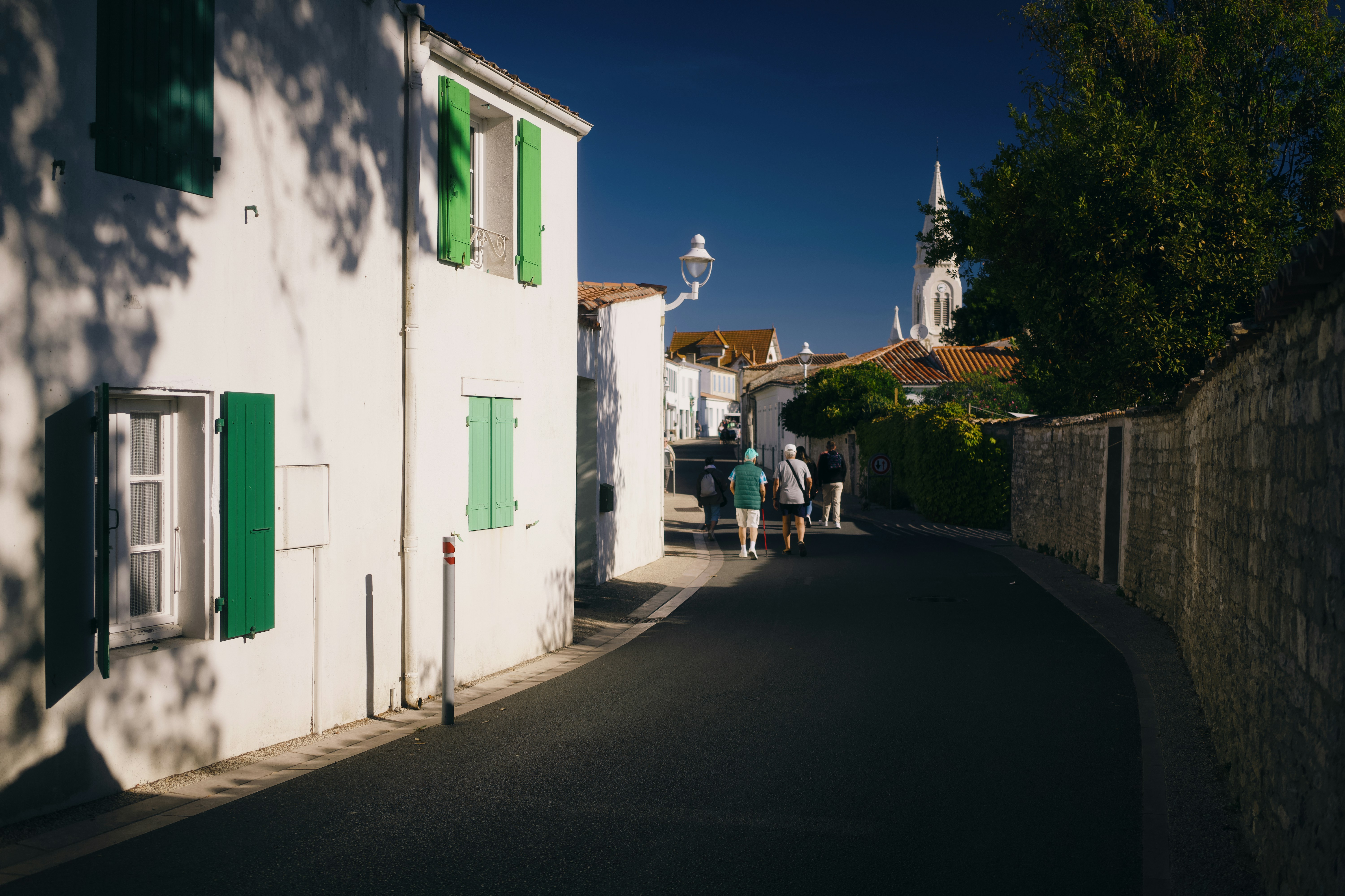 People walk down a sunny, narrow street with white buildings.