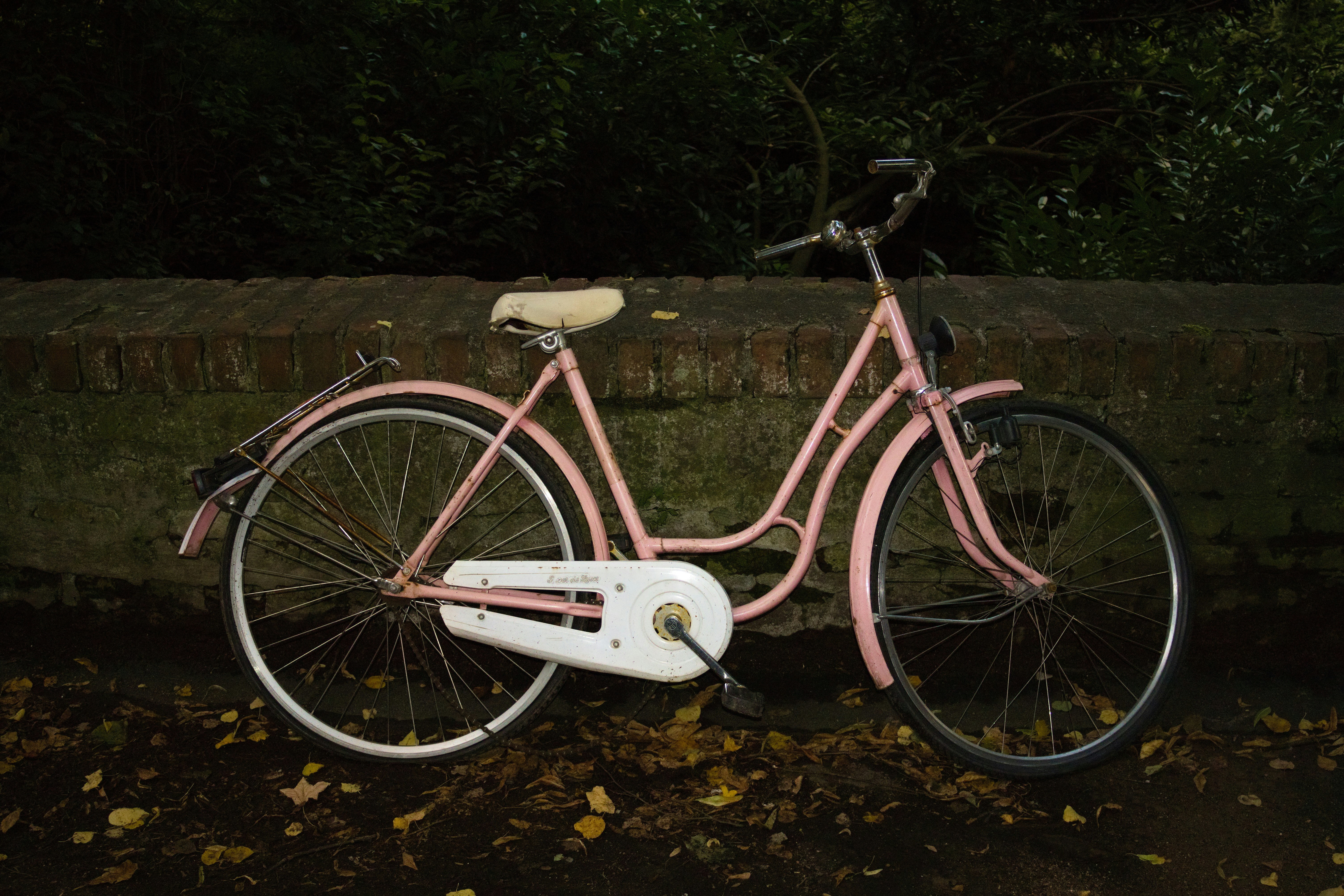 An old bike leaning against a brick wall on a small bridge. | A pink bicycle parked by a brick wall