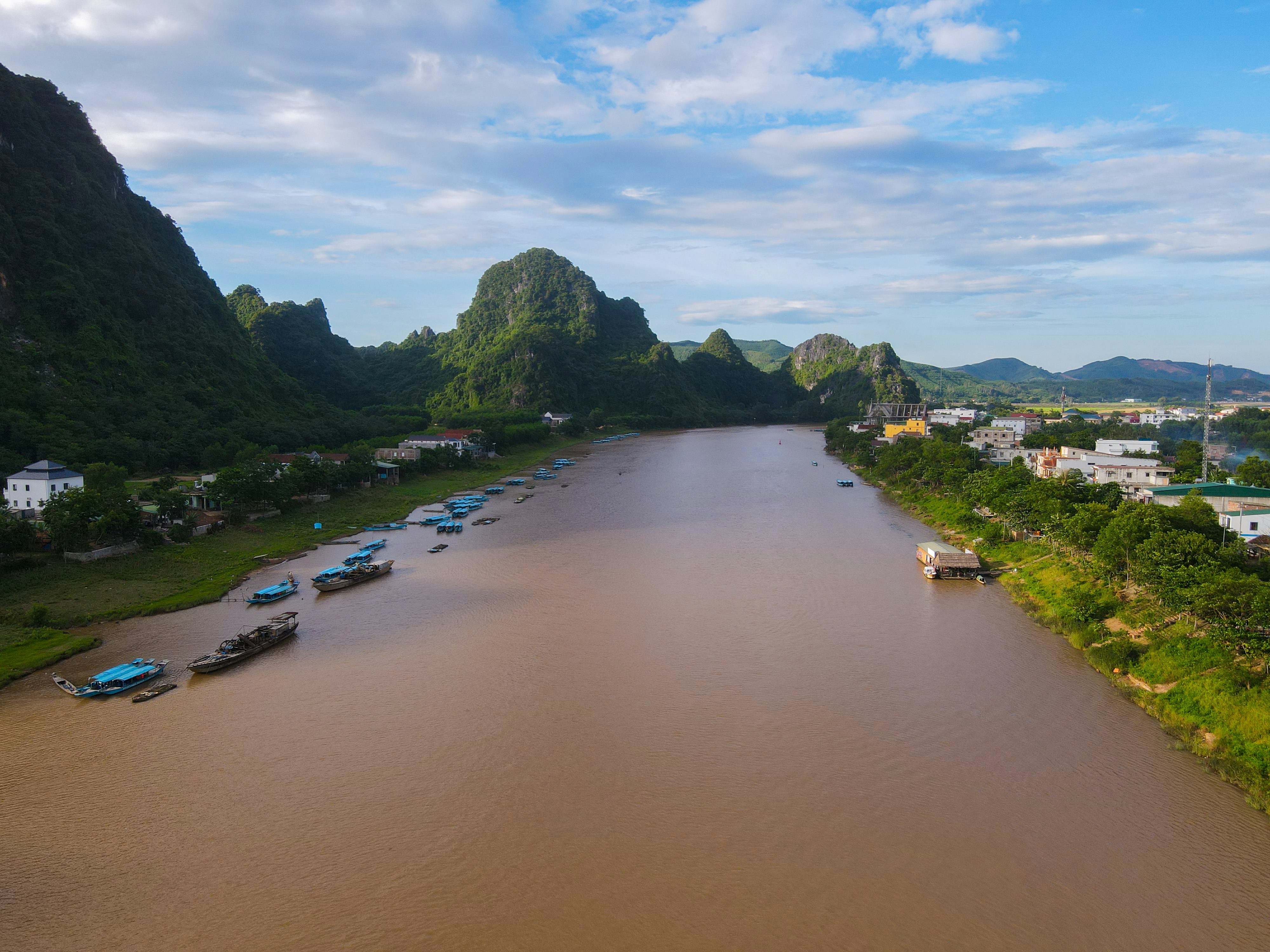 Aerial photograph of a muddy river winding between lush karst hills, with boats docking along the banks and a distant settlement.