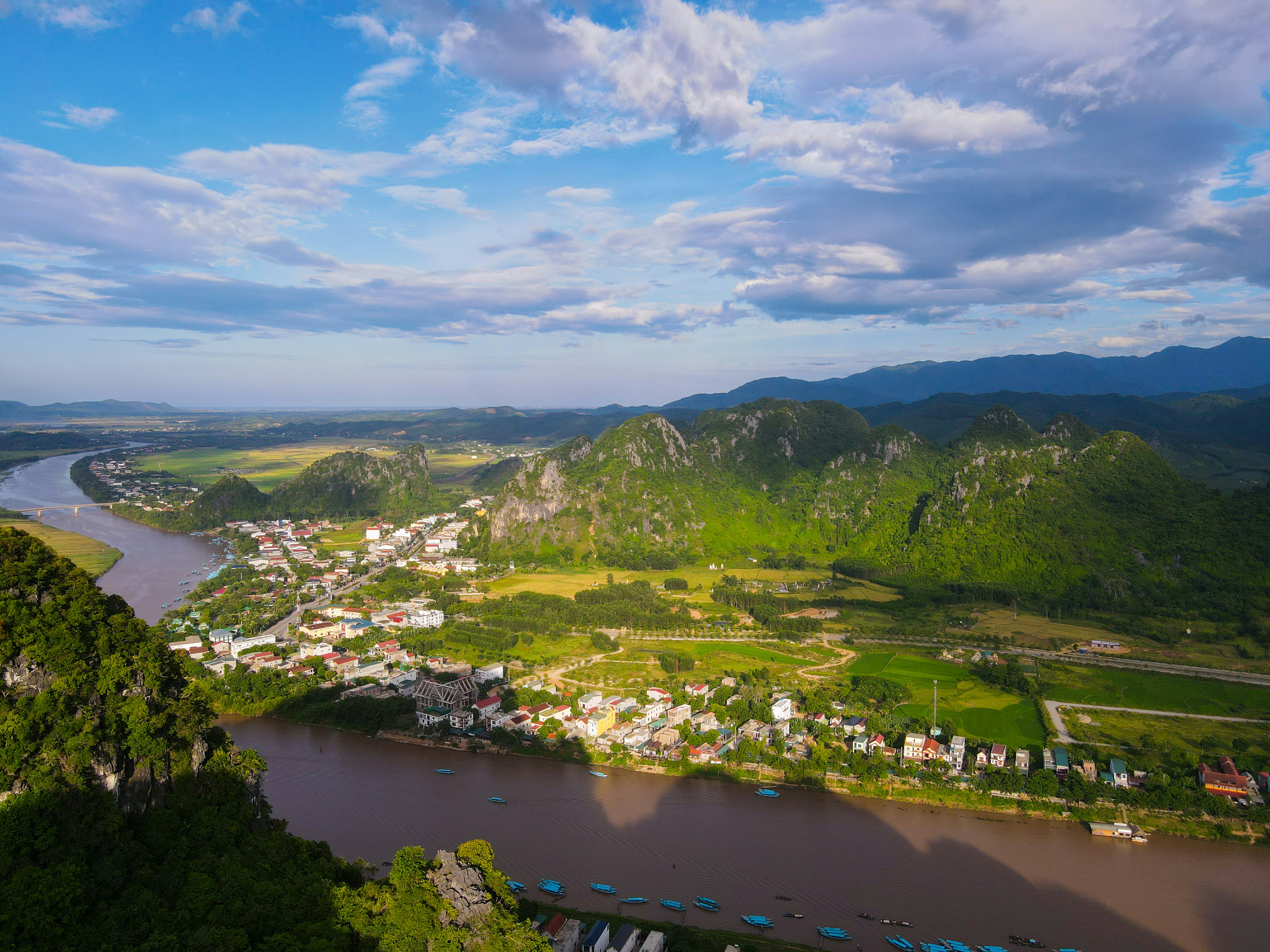 Aerial view of a lush river valley with a quaint village nestled against verdant hills, showcasing the interplay of land and water under a vibrant sky.