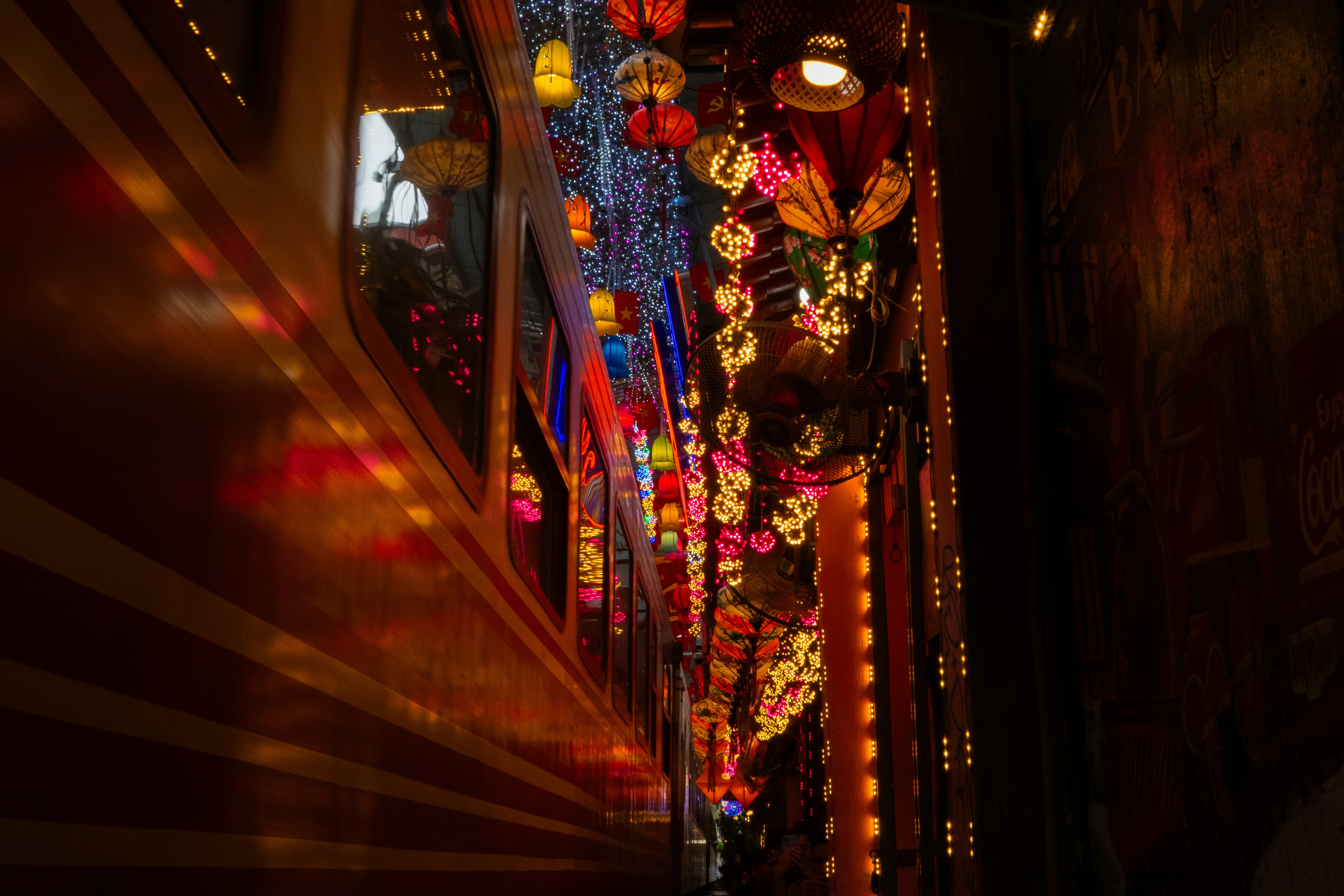 Vibrant alleyway adorned with colorful lanterns and lights, reflecting off a vintage train. The atmosphere is festive and lively.