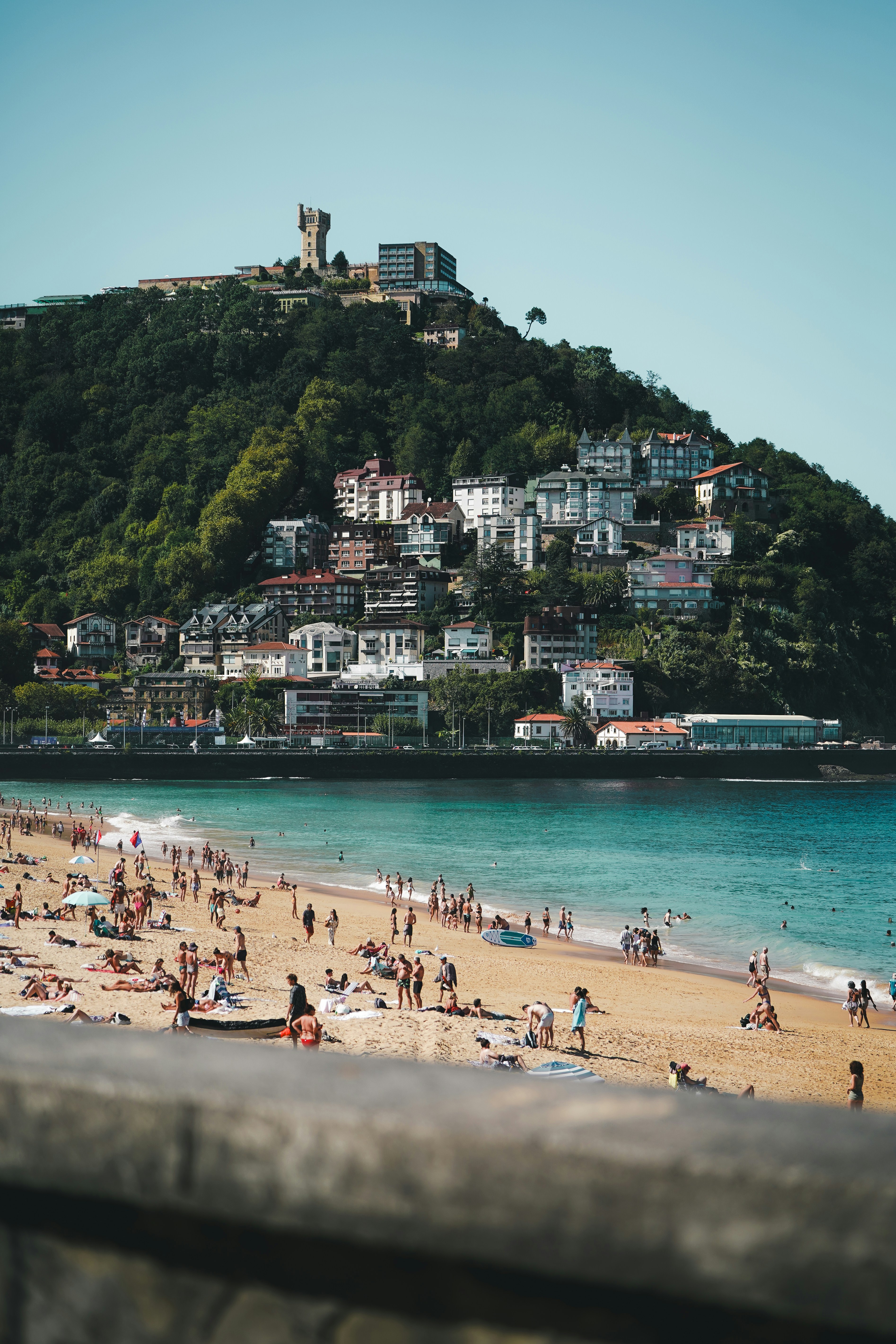 Crowded beach with buildings on a green hillside.