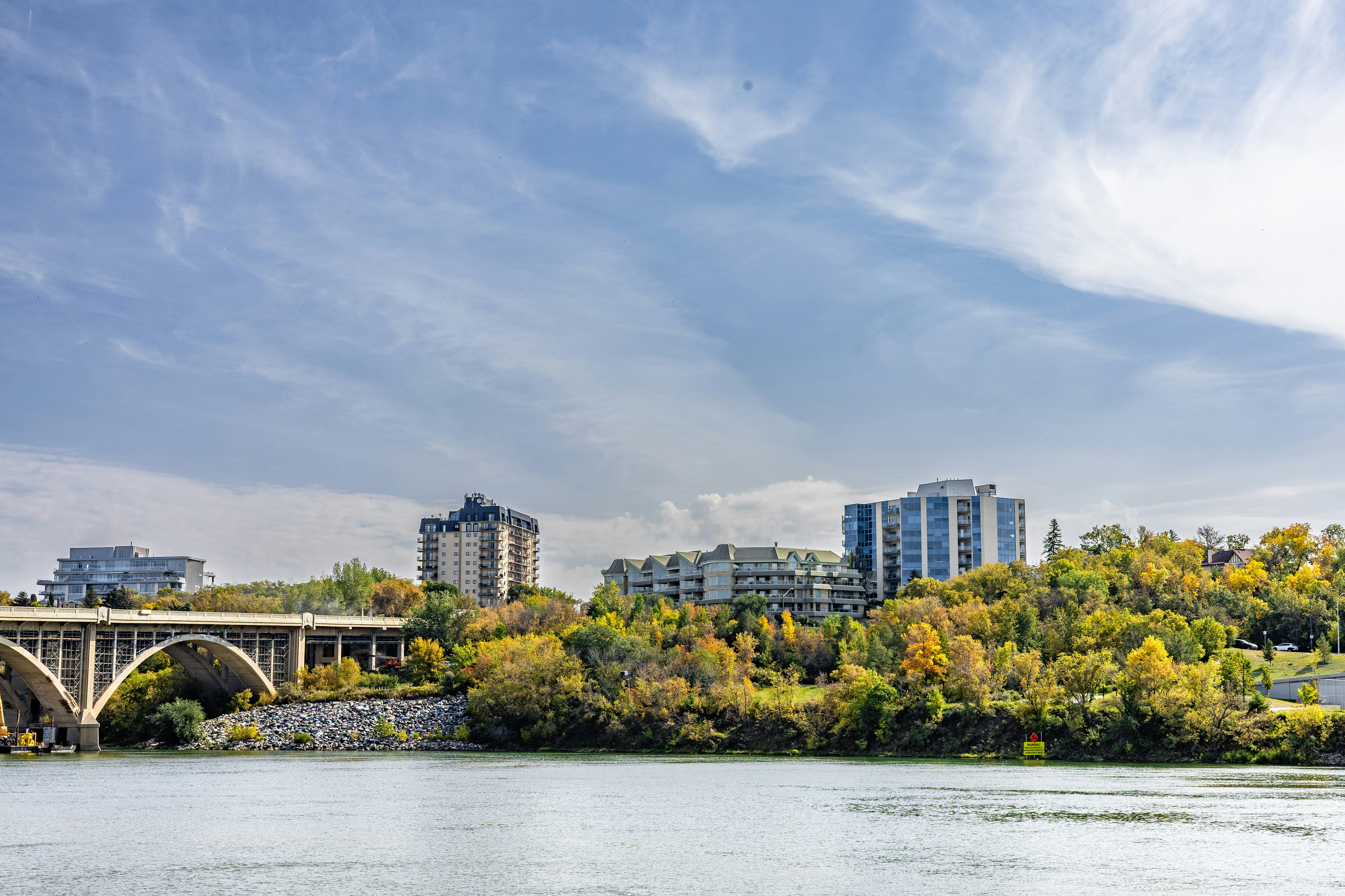 Vibrant autumn foliage frames modern buildings along a serene riverbank under a dynamic sky.