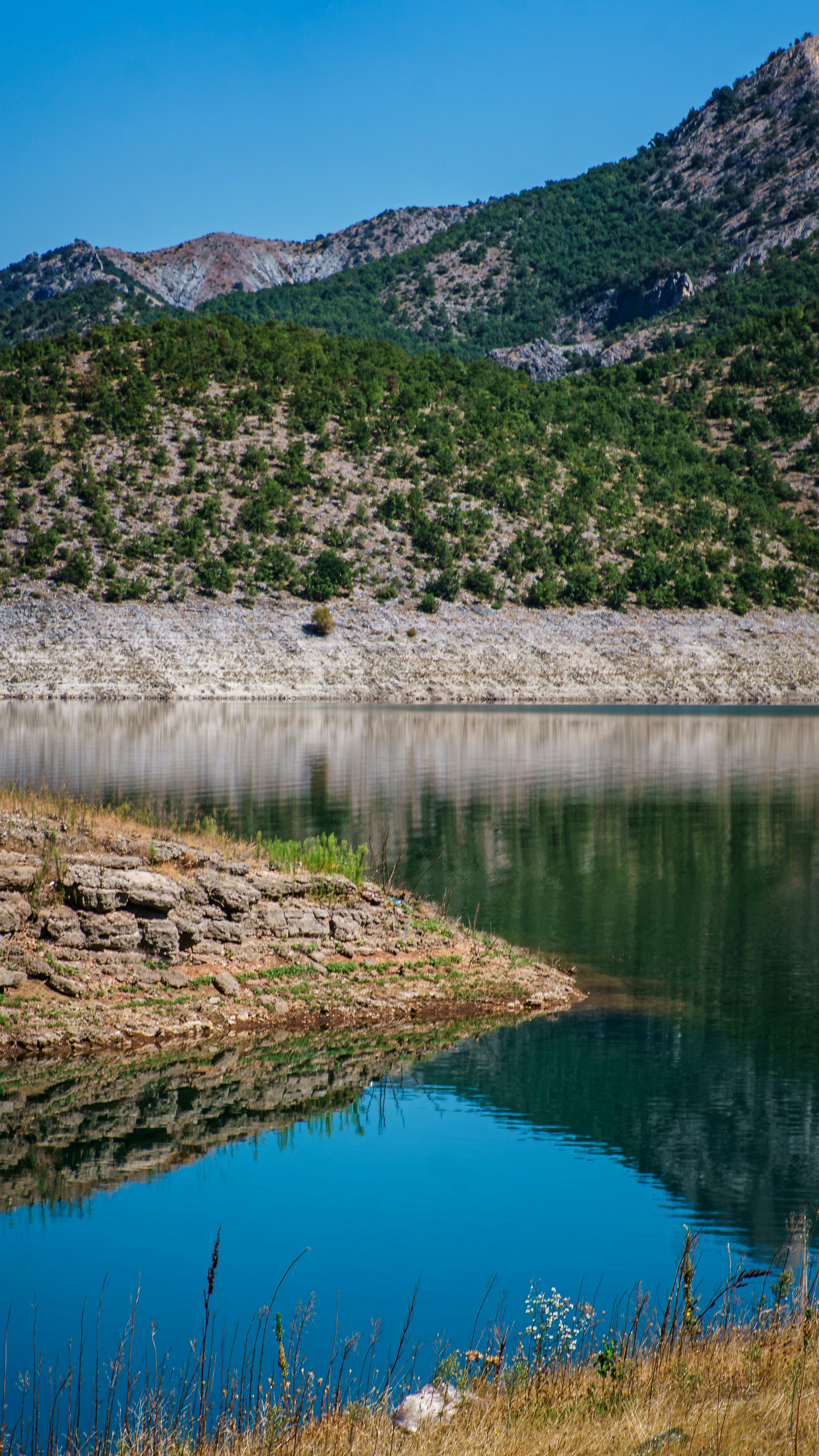 Calm lake reflecting the surrounding mountains and greenery under a clear blue sky.