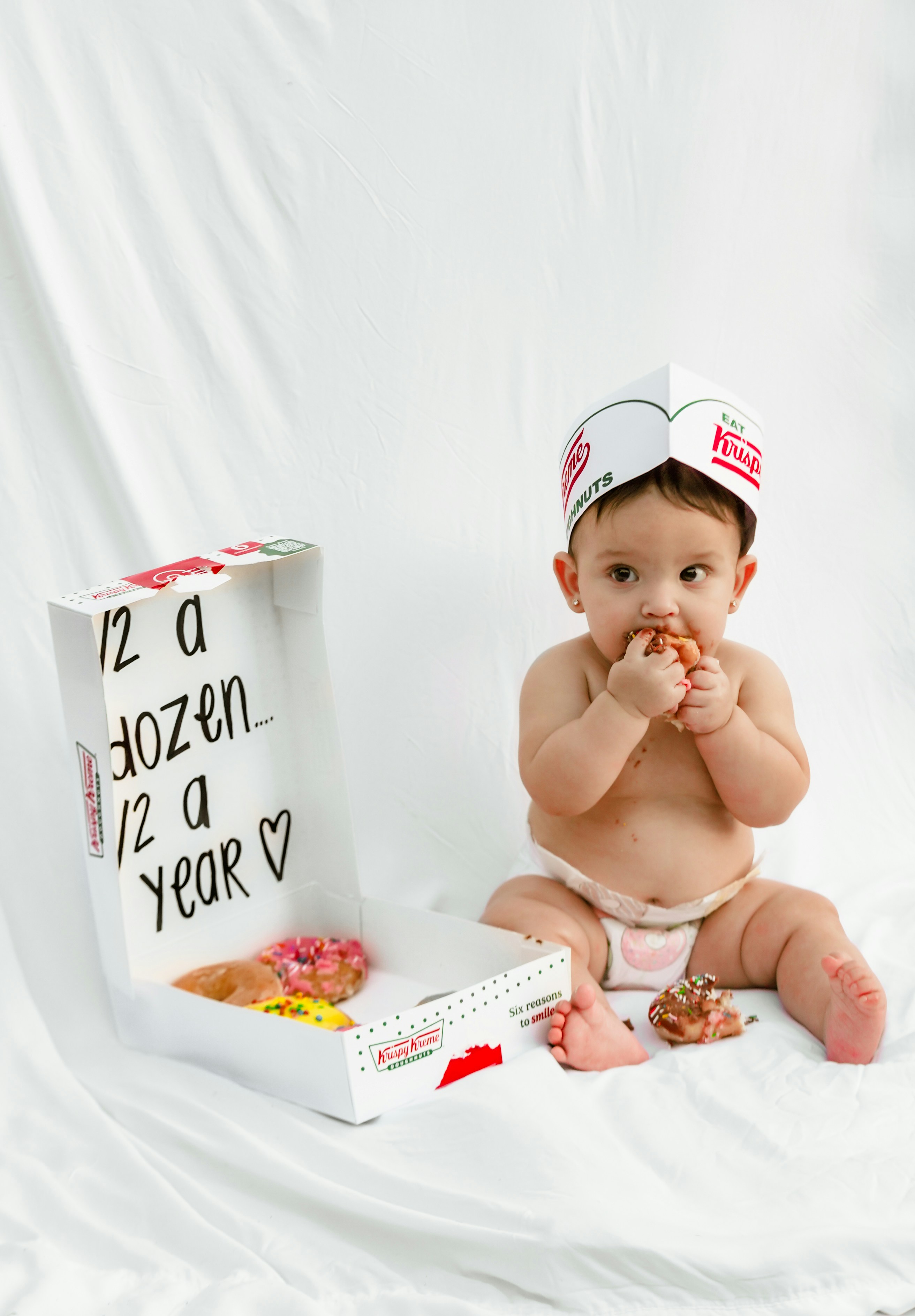 A toddler in a Krispy Kreme hat enjoys a donut while seated next to an open box of assorted donuts, embodying pure joy and innocence.