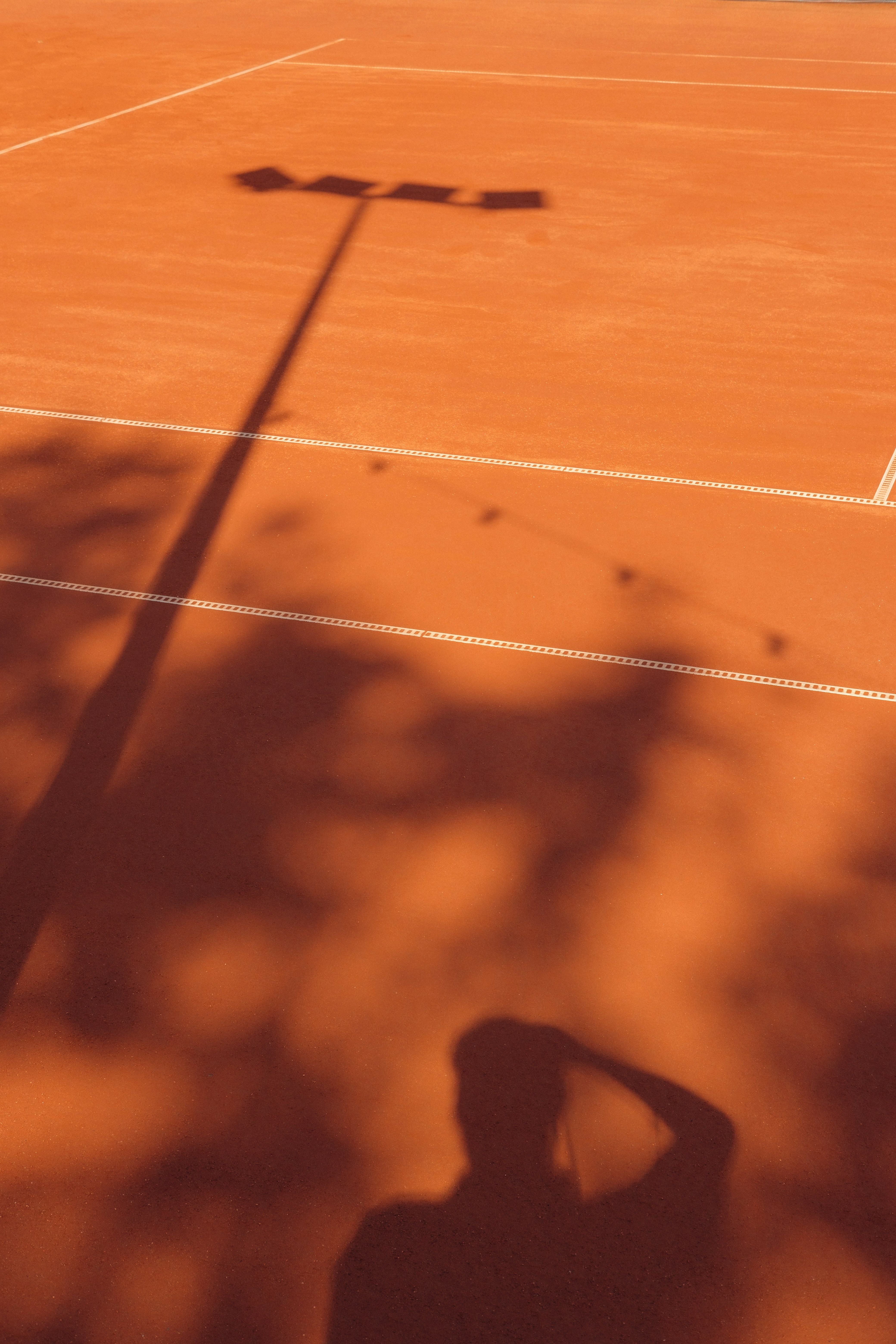 A shadow cast by a lamp on a clay tennis court, with faint lines marking the boundaries. The warm tones evoke a sense of quiet anticipation.