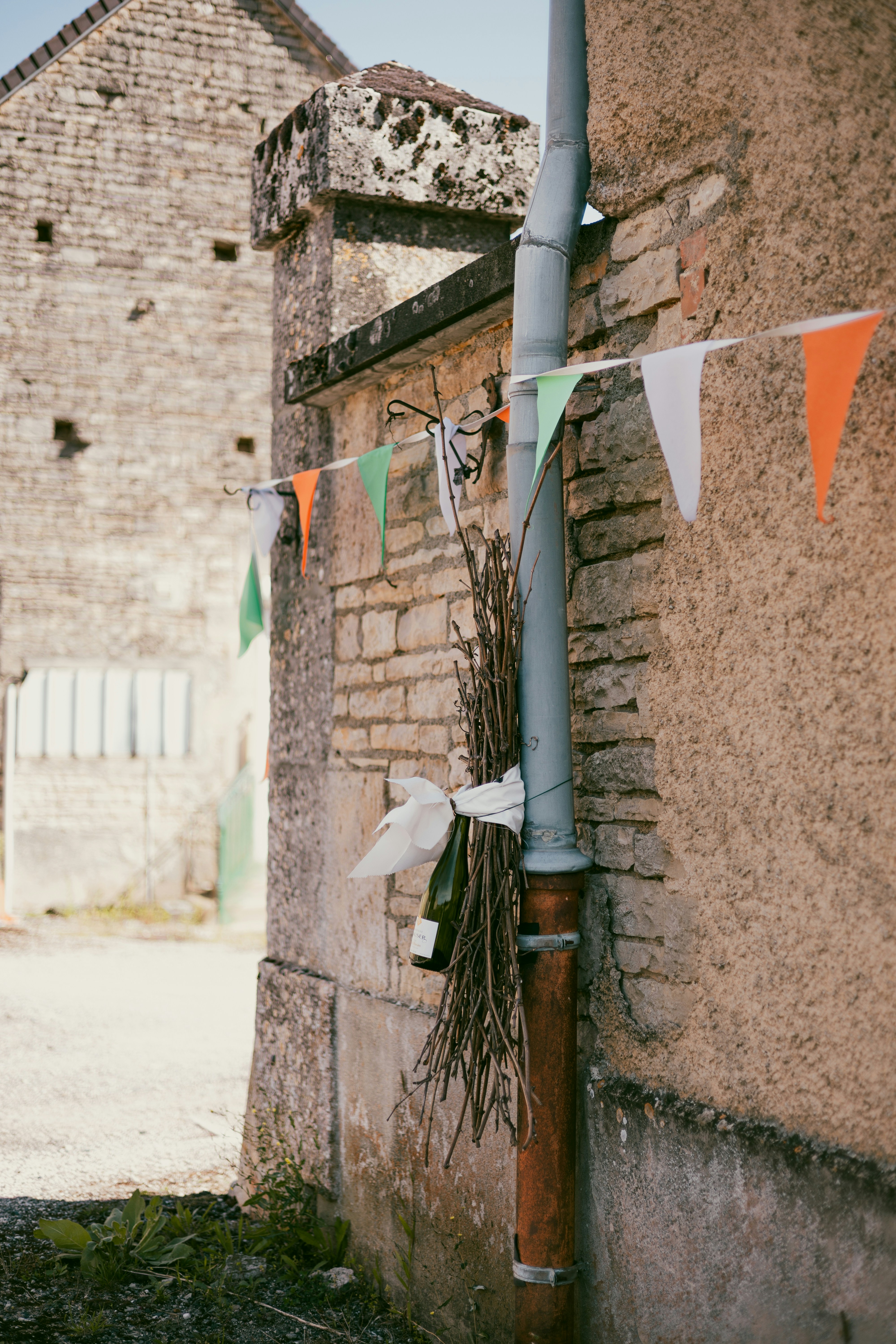 Twig wreath on a rusted pipe under pastel bunting in a sunlit stone alley
