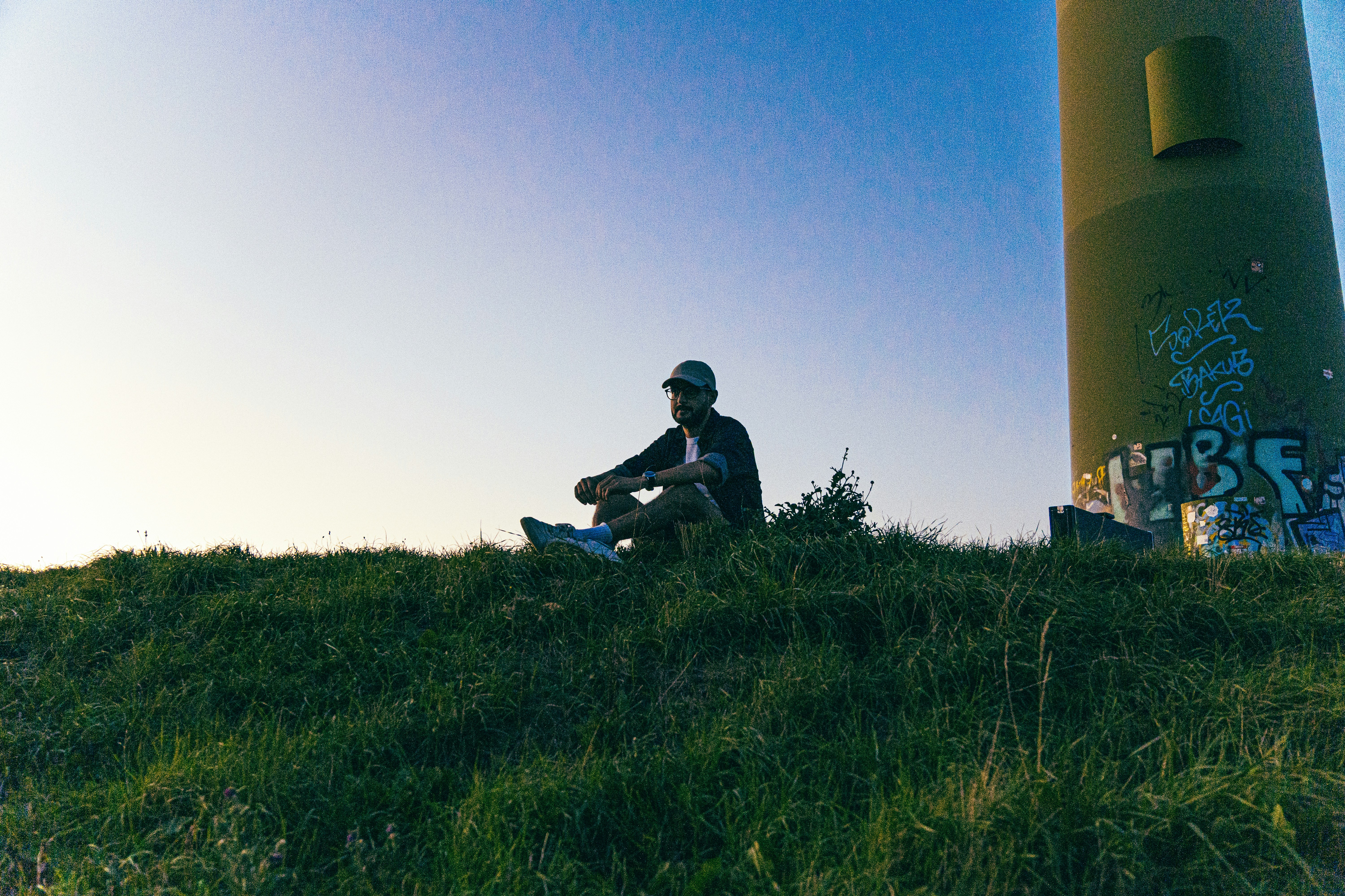 A solitary figure sits on a grassy knoll, gazing thoughtfully at a colorful graffiti-covered structure under a vast sky.