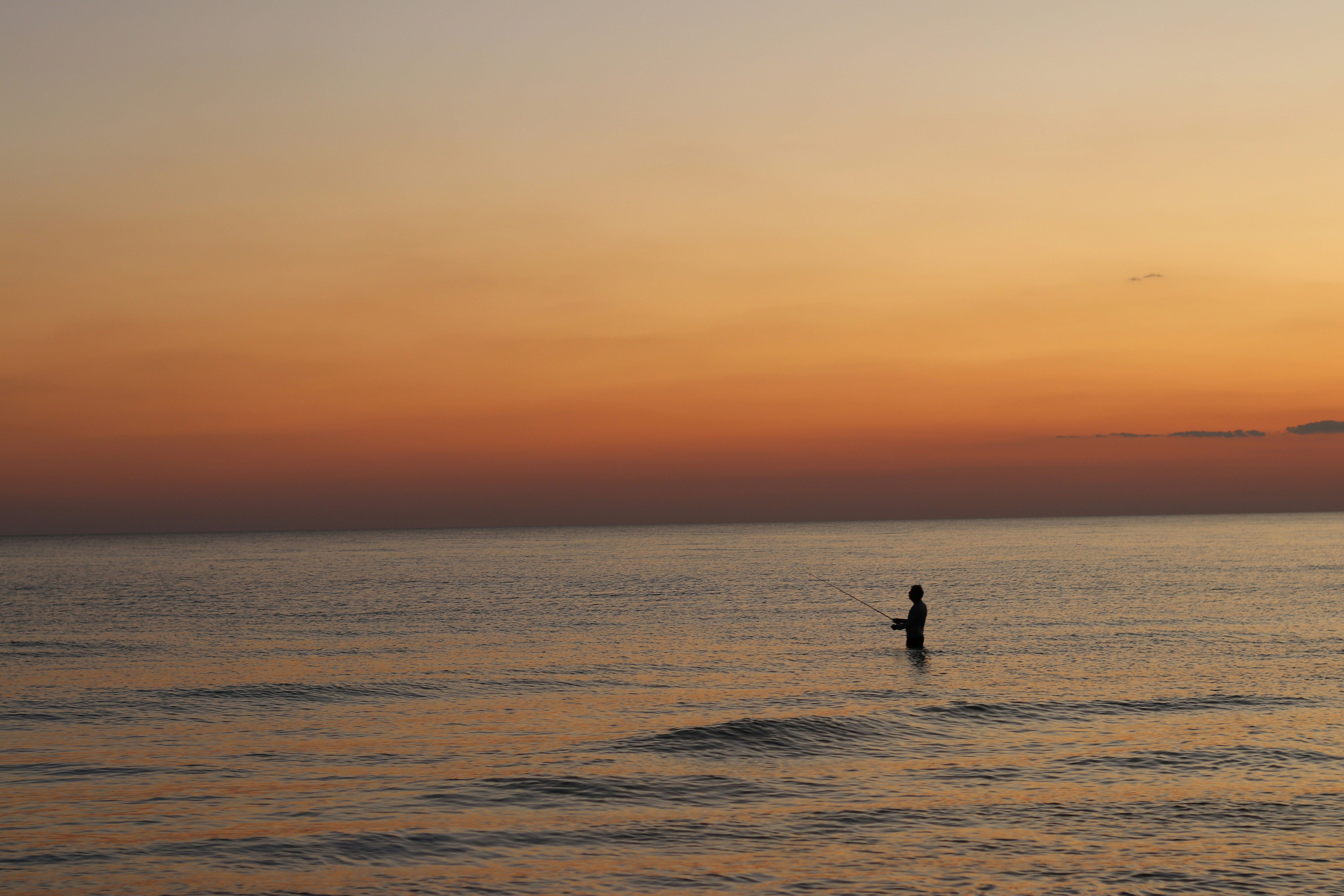 A lone fisherman stands in calm waters, silhouetted against a vibrant sunset sky with hues of orange and purple.