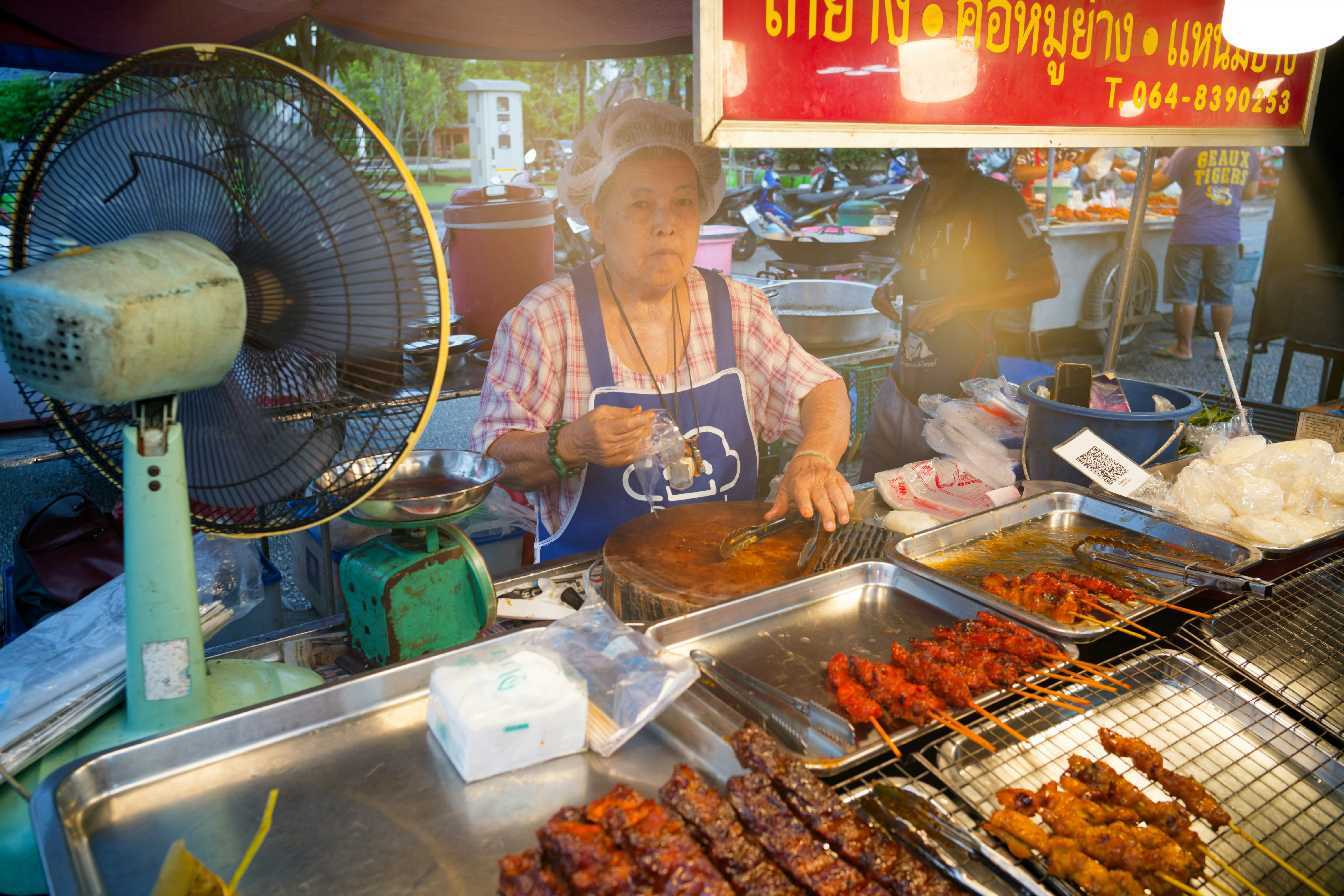 A street vendor skillfully preparing skewered meats at a bustling market, surrounded by an array of delicious offerings. The vibrant atmosphere is enhanced by the presence of a cooling fan.