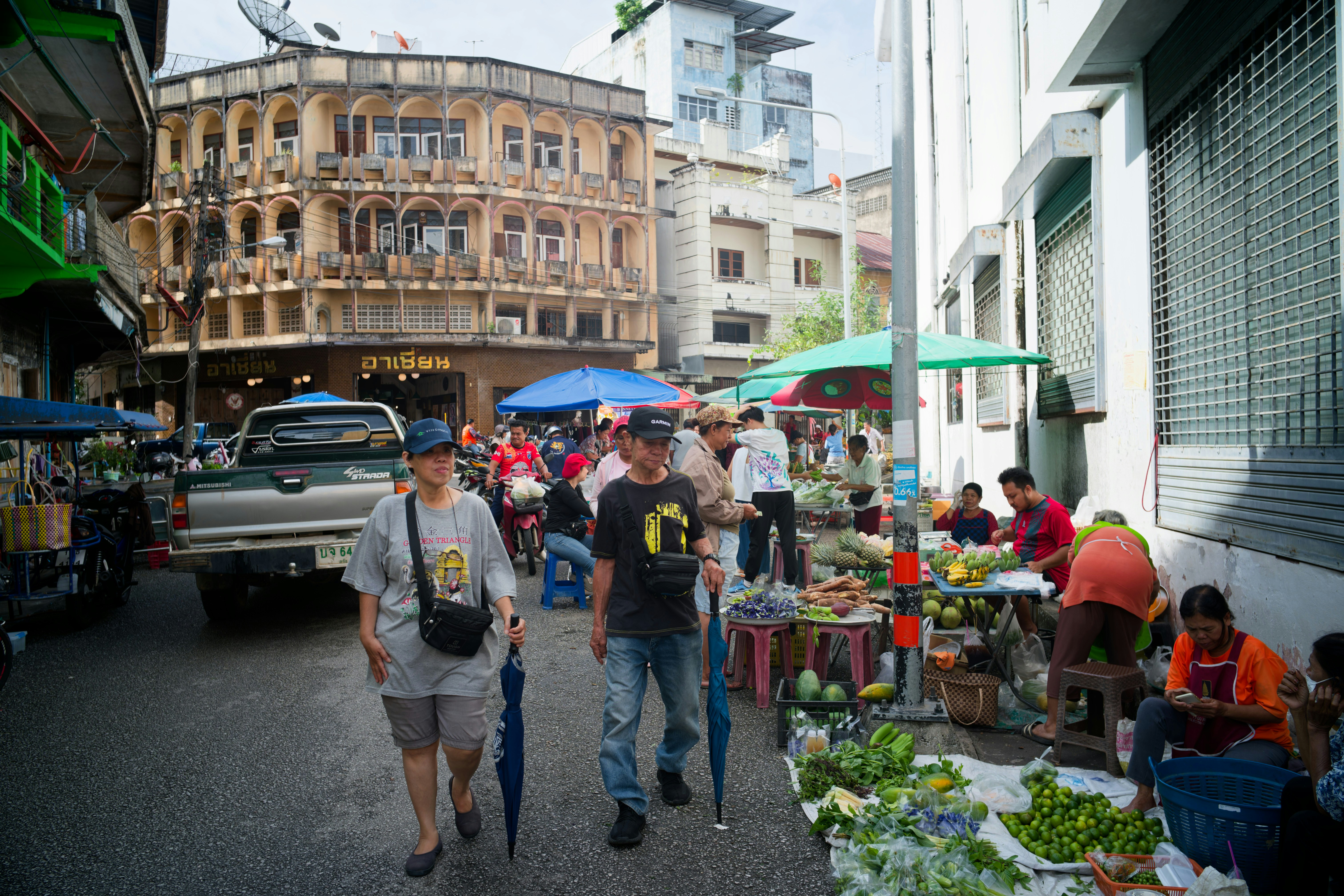 Bustling street market scene with vendors selling fresh produce and shoppers navigating colorful umbrellas. A vintage building looms in the background.