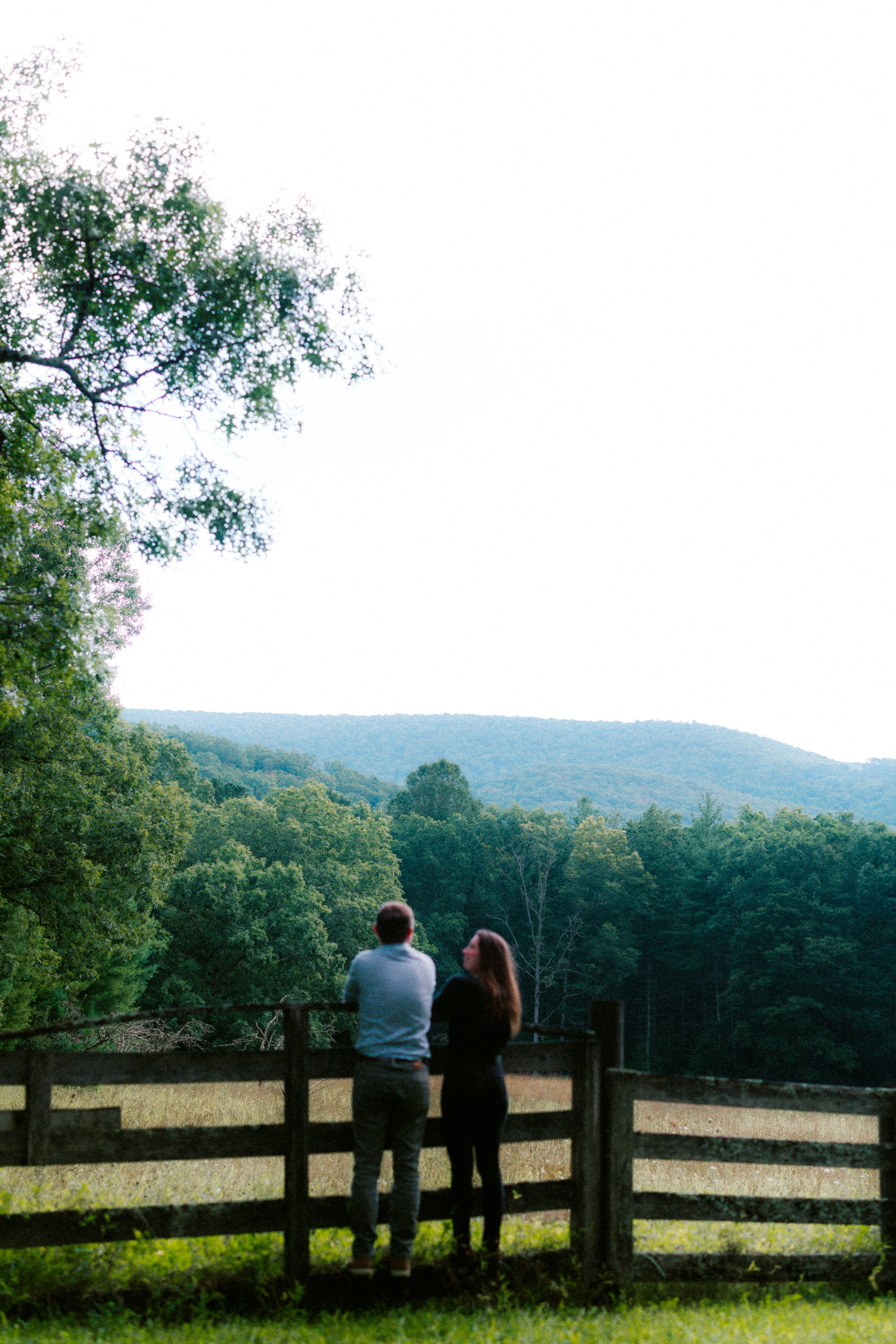 Couple standing by a wooden fence, gazing at expansive green hills under a soft sky. The serene landscape invites contemplation.