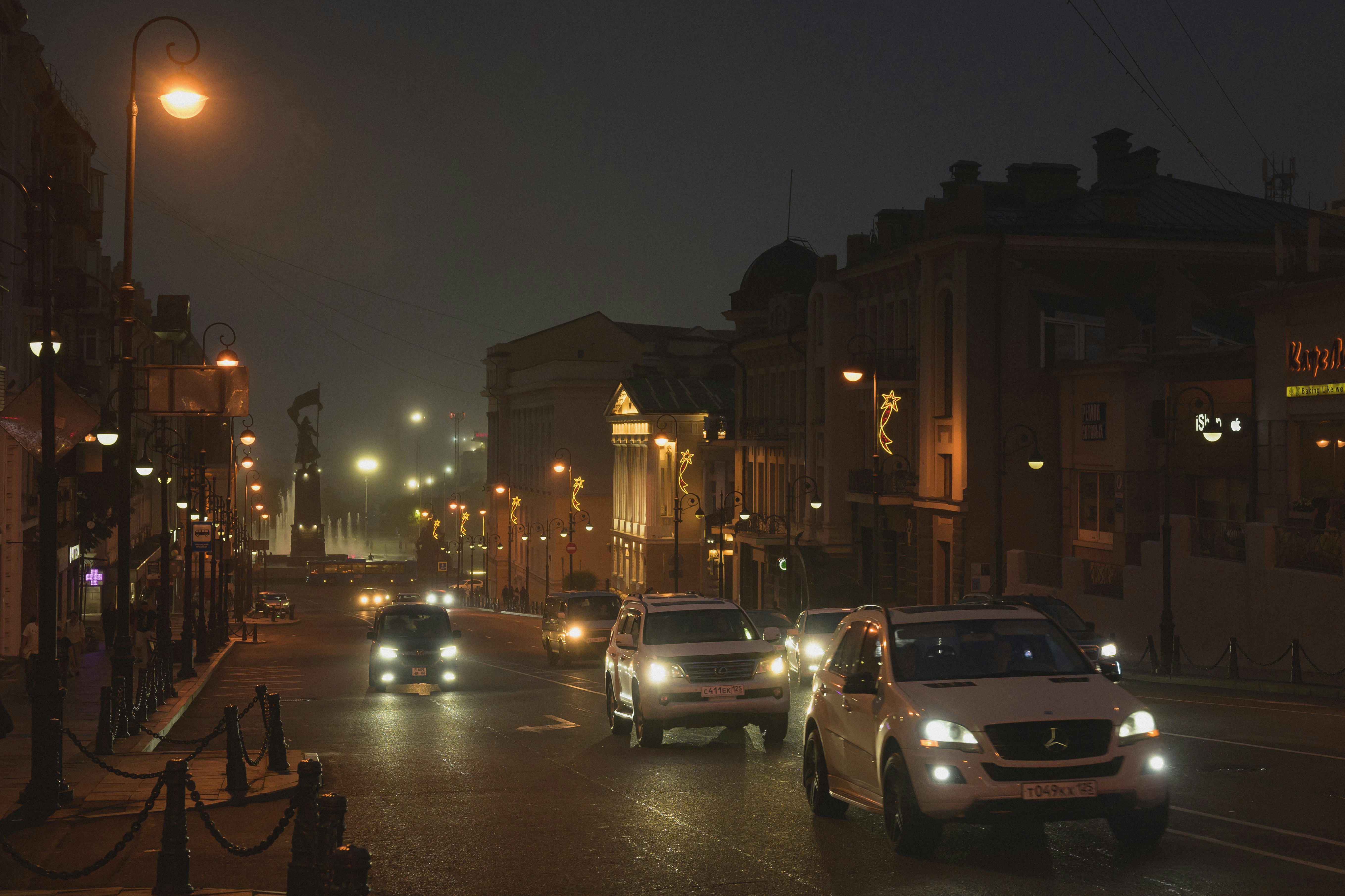 A moody evening street scene in Vladivostok, Russia, with glowing streetlights reflecting off the wet pavement. Historic buildings line both sides of the road, while headlights cut through the mist. In the distance, the Monument to the Fighters for Soviet Power stands tall, anchoring the city’s dramatic nightscape. | Cars driving on a wet city street at night.