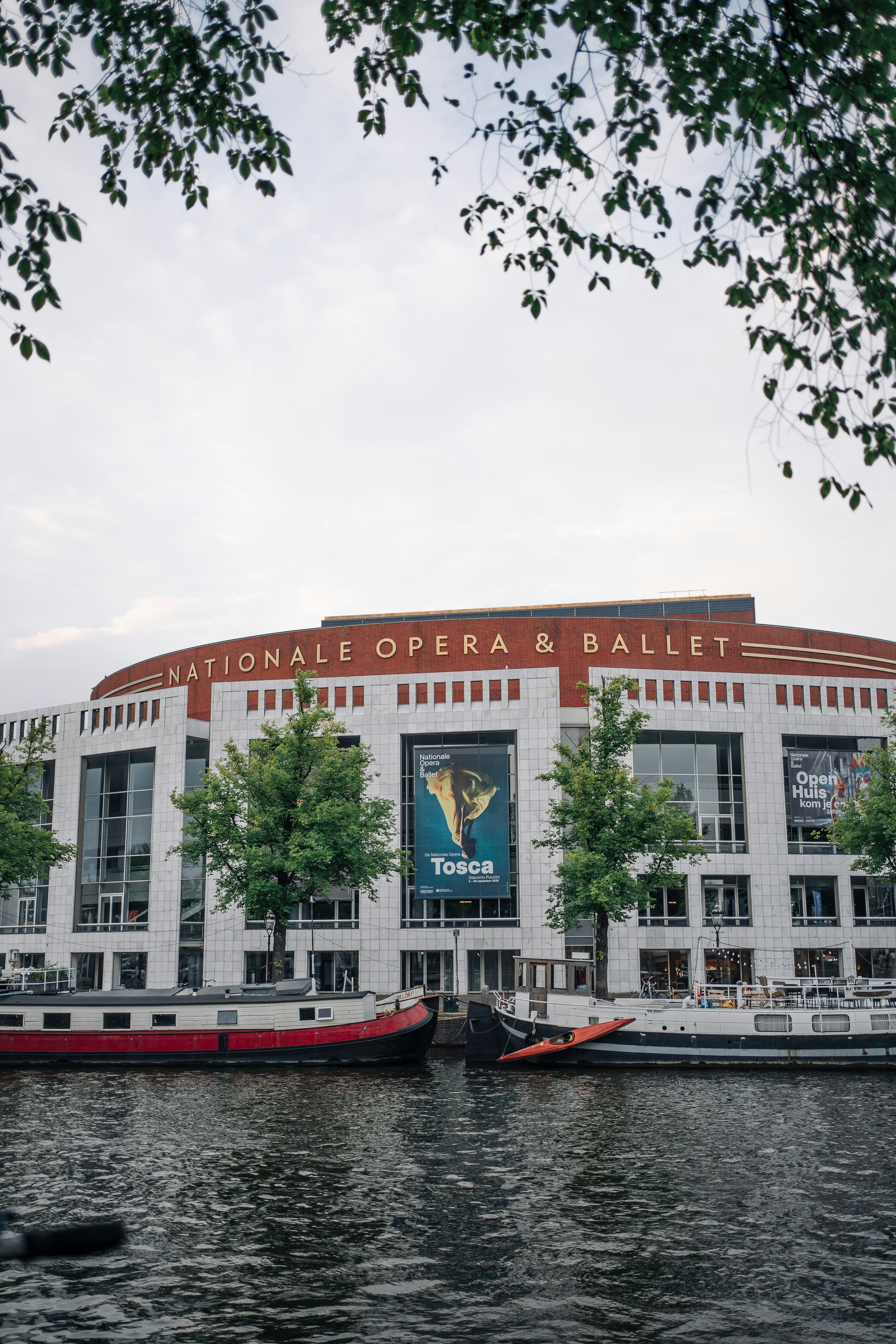 Modern opera house featuring a prominent poster for Tosca, framed by lush trees and a canal view.