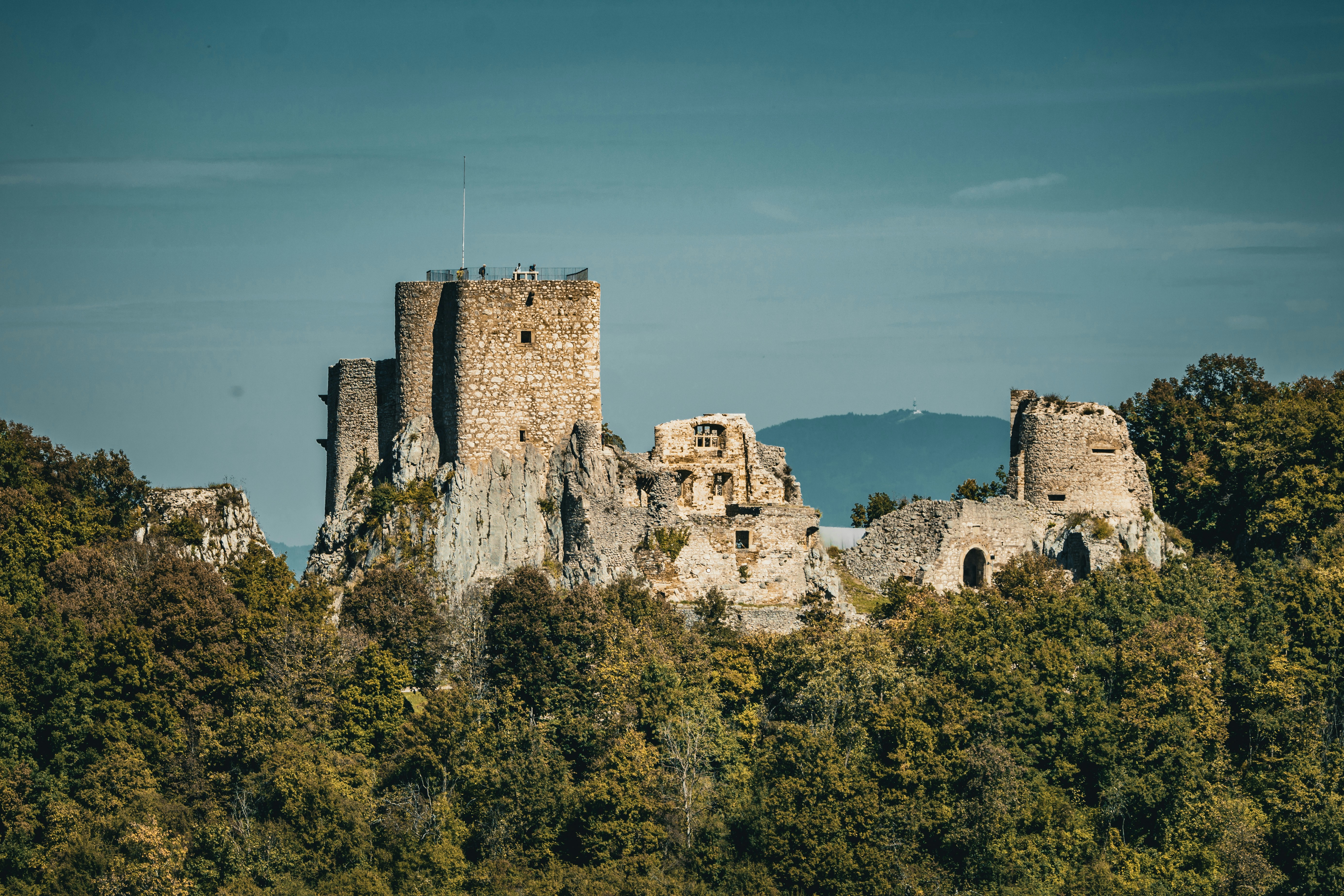 Perched atop a rocky hill and surrounded by dense forest, the weathered ruins of a medieval stone castle stand proudly against a clear sky, echoing centuries of history and forgotten battles. Its crumbling walls and enduring tower evoke mystery and medieval grandeur. | Ancient stone castle ruins on a forested hill.