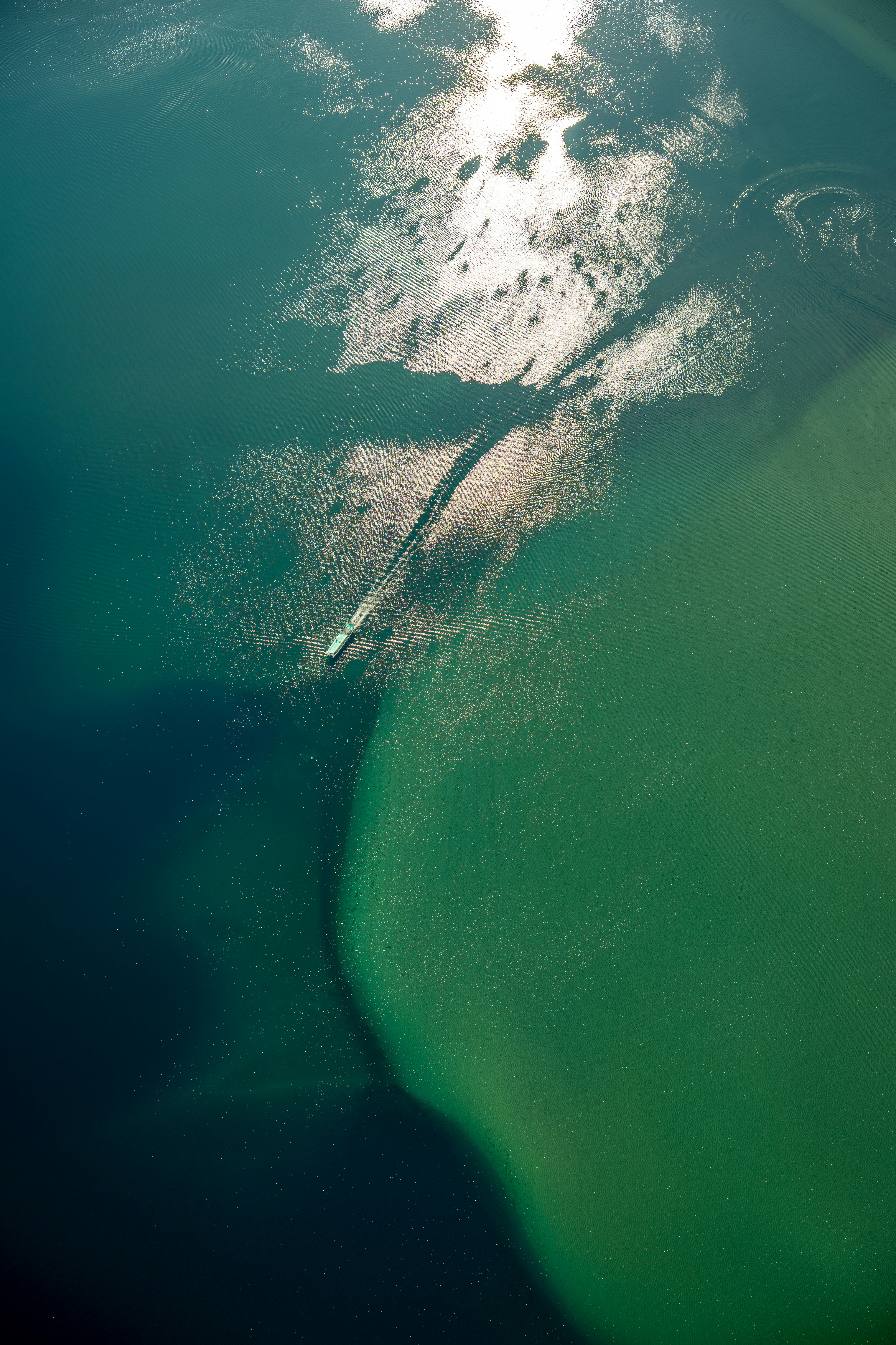 Aerial view of a boat navigating through shimmering emerald waters, with sunlight creating sparkling reflections on the surface.