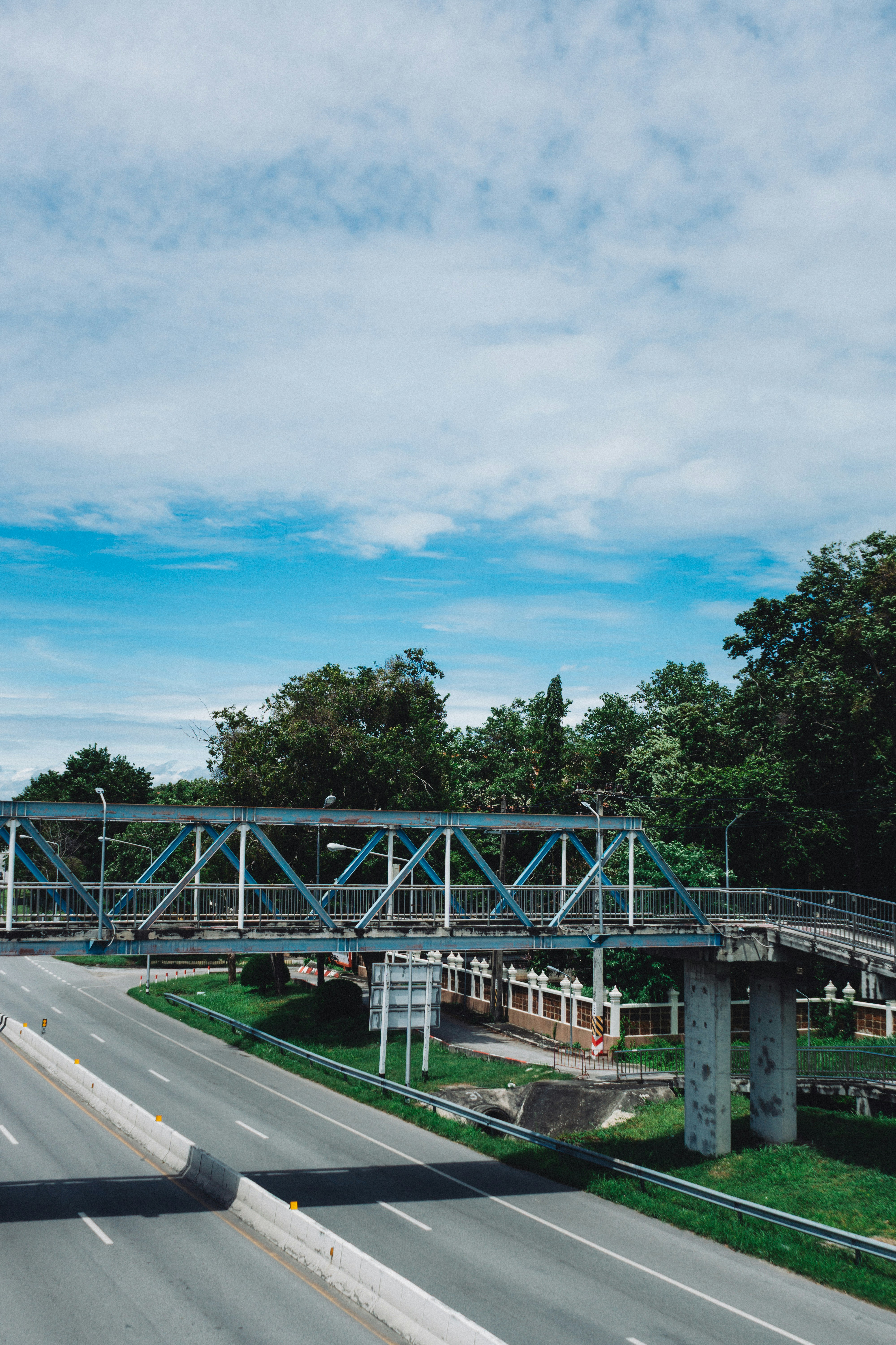 A pedestrian bridge crosses over a busy road, surrounded by lush greenery and a bright blue sky.