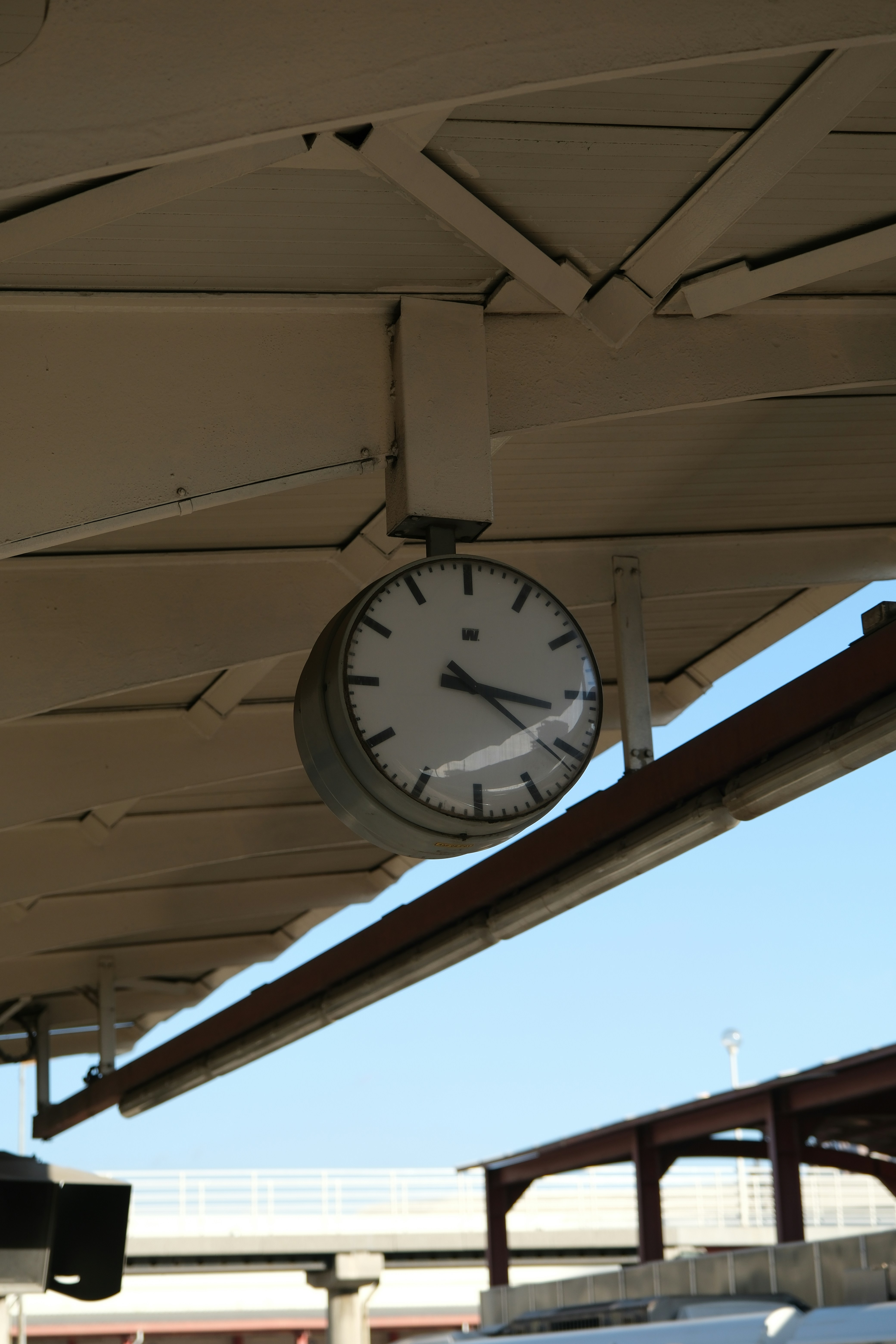 A hanging station clock captured from a low angle, showing the time as 10:20, with the station roof structure above and clear sky in the background. | A round clock hangs under a canopy.