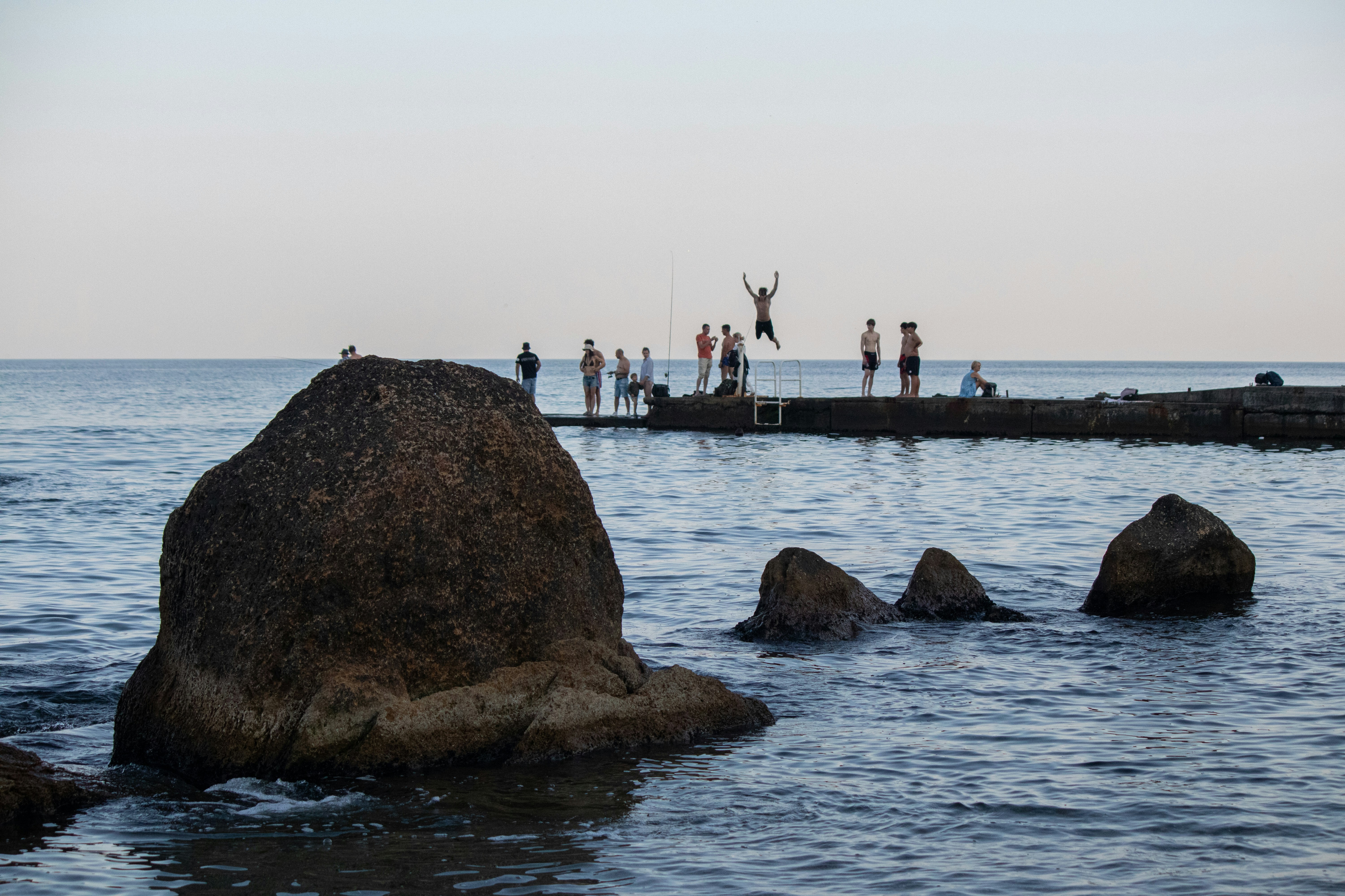 A group of people enjoying a summer evening by the water, with one individual leaping off a pier into the sea. Large rocks dot the shoreline.