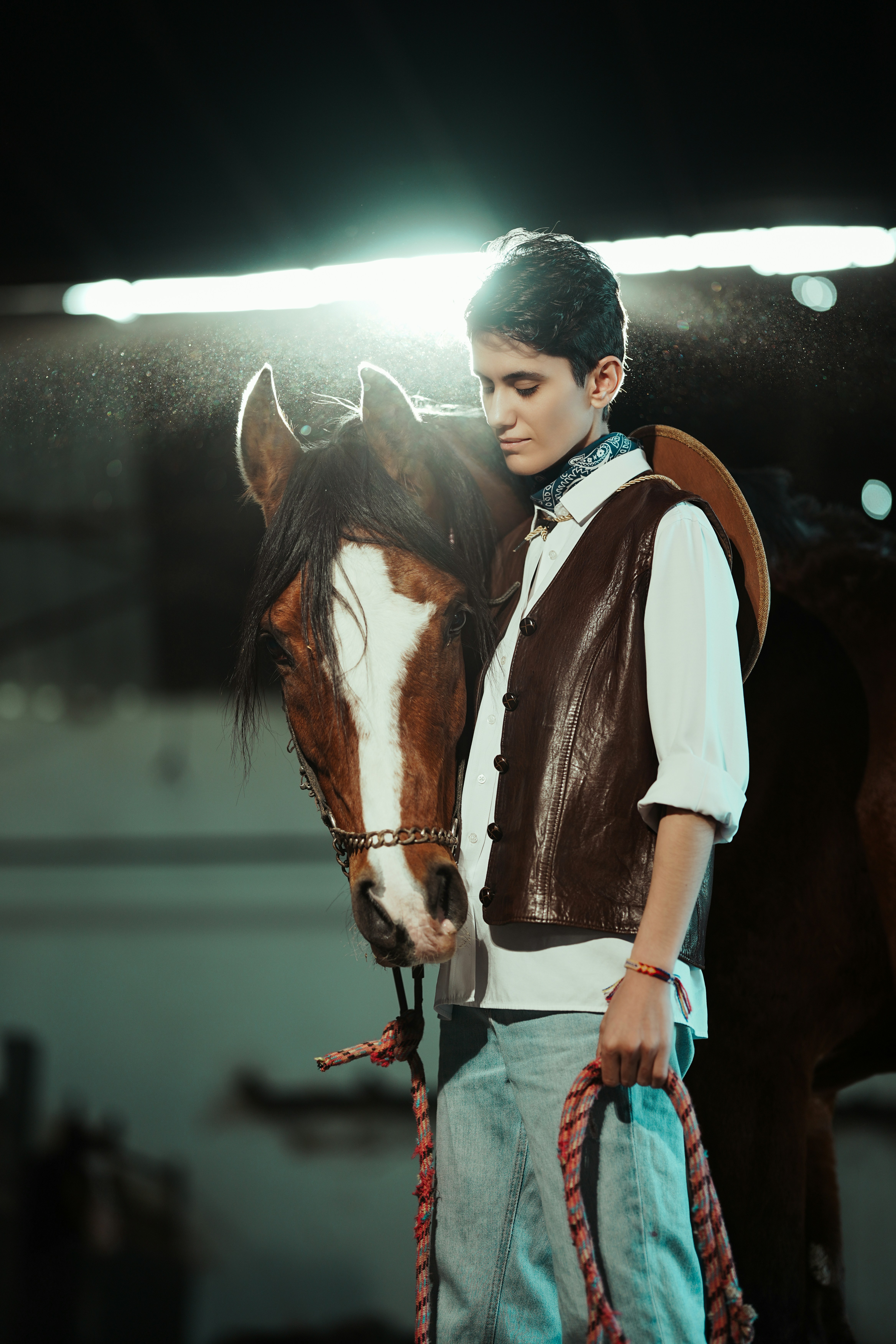 A young cowboy gently embraces a horse, showcasing the bond between them in a softly lit stable environment.