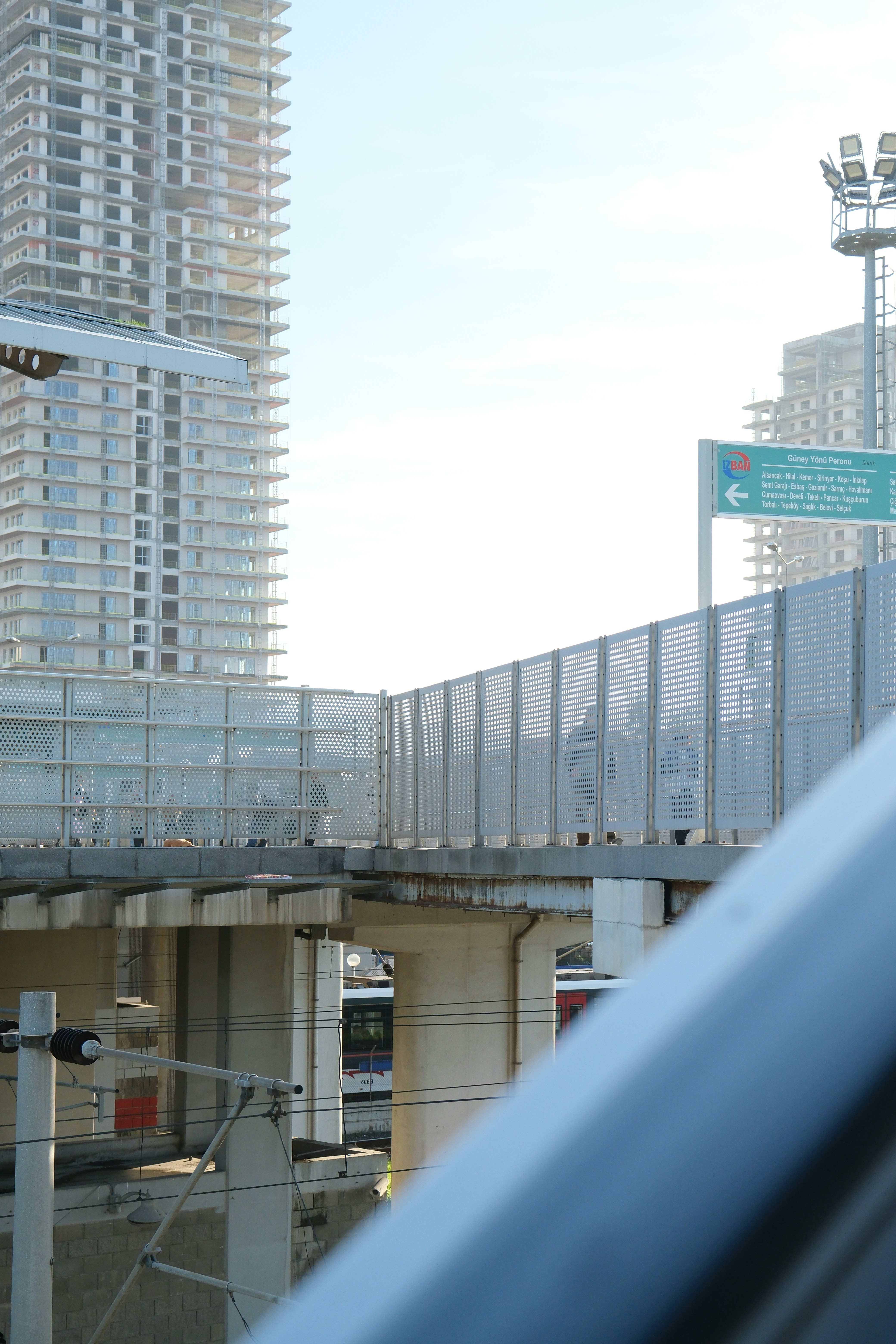 Urban scene featuring high-rise residential buildings in the background and an elevated pedestrian bridge with metallic fencing in the foreground. The composition highlights modern city infrastructure with a mix of architecture, signage, and transport elements.
