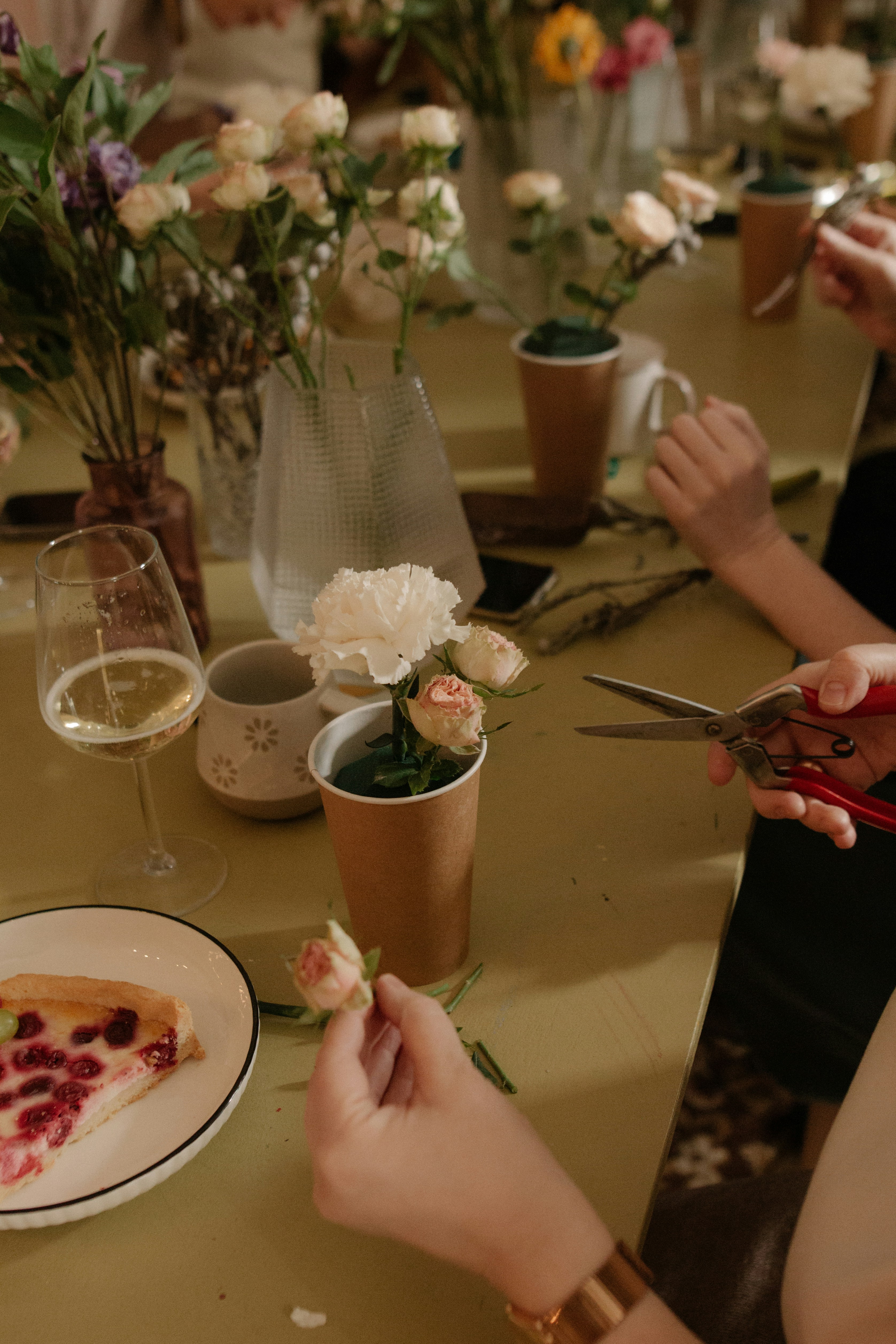 Hands delicately trimming flowers at a floral arrangement workshop, surrounded by vases and refreshments. A slice of pie rests on the table, adding a cozy touch.