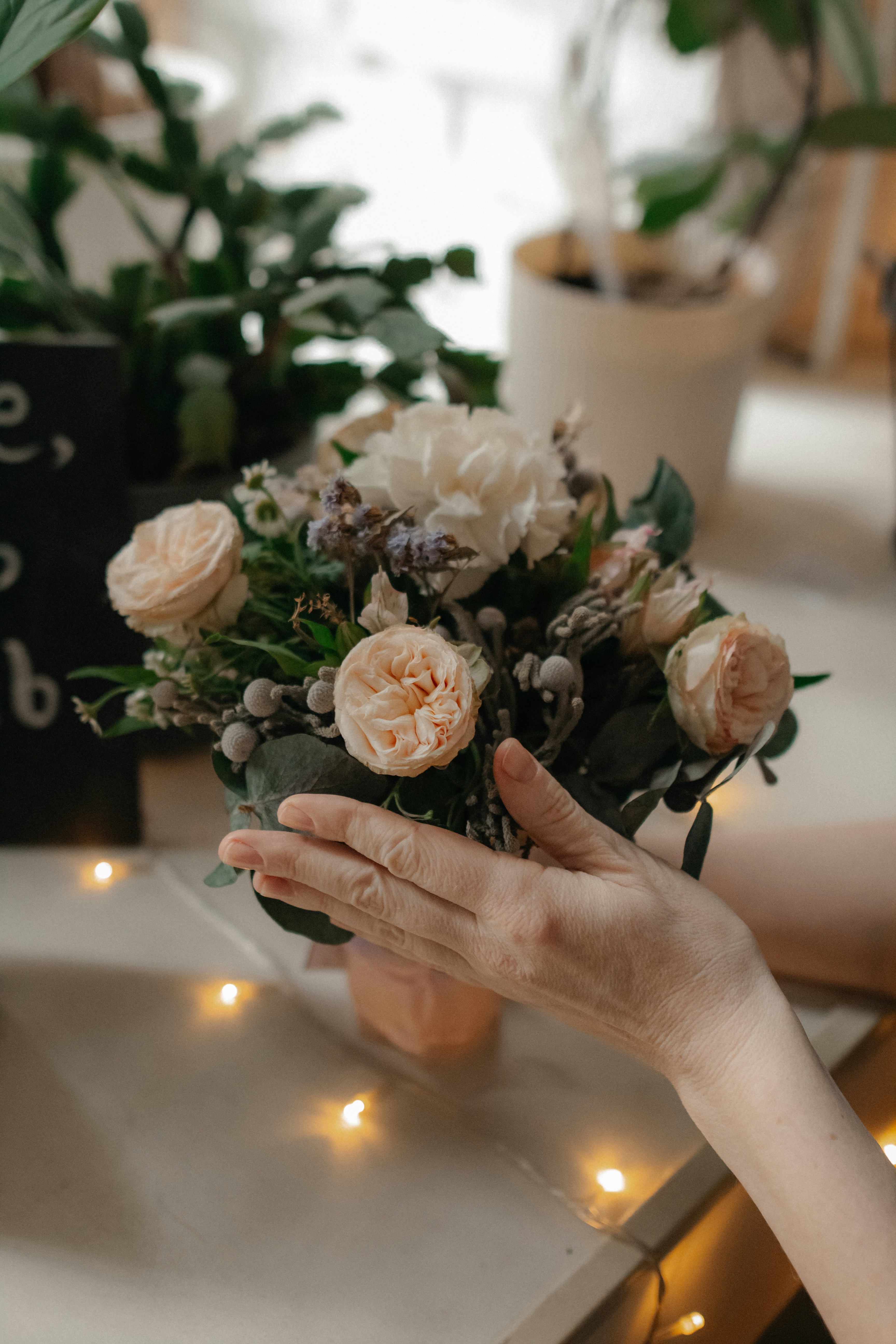 A hand gently cradles a bouquet of soft-hued flowers, surrounded by lush greenery and warm ambient lights.