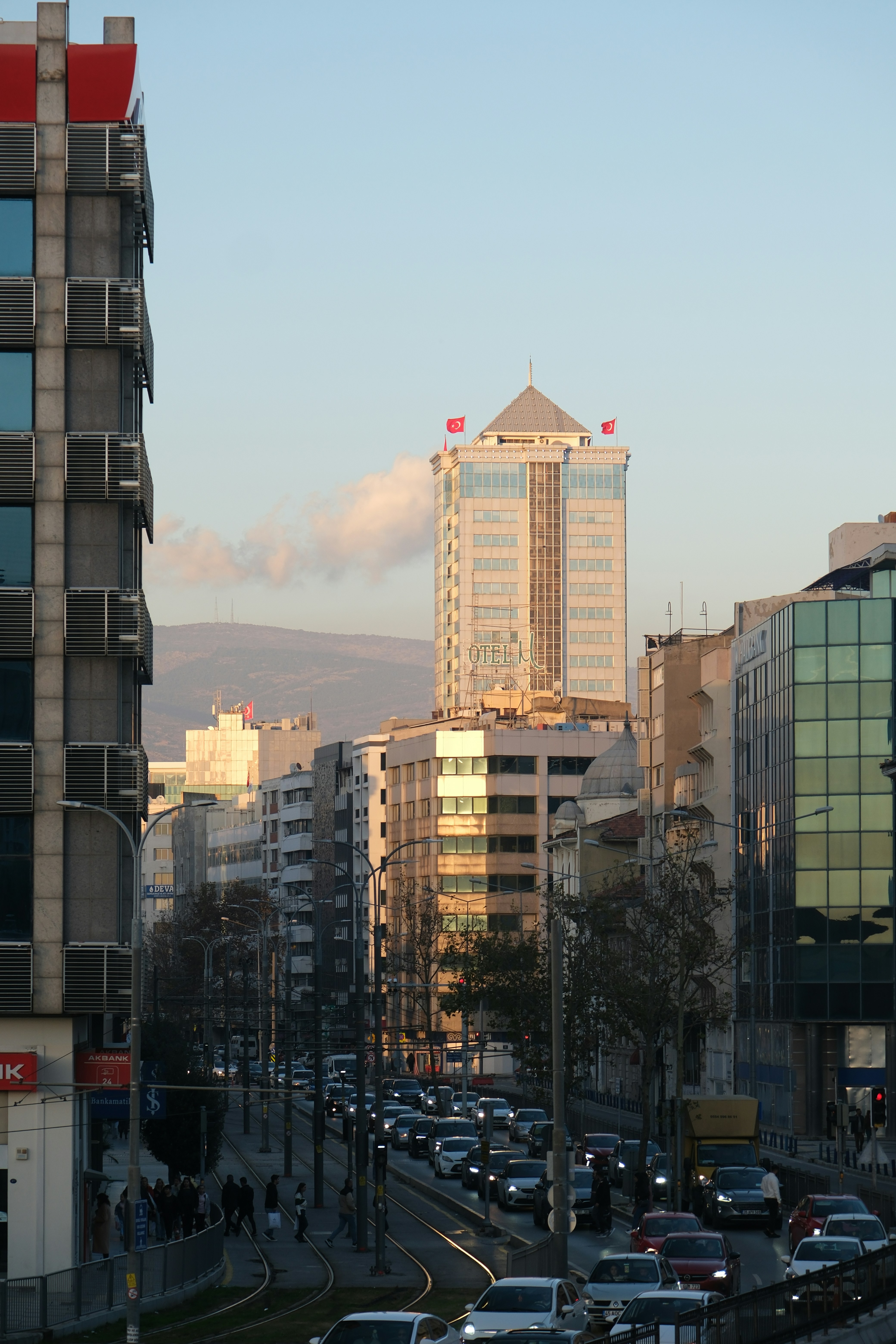 A cityscape at sunset featuring tall modern buildings, one of them crowned with Turkish flags. The scene captures a busy avenue filled with traffic and tram lines, while the warm evening light reflects off the glass facades. | City street with traffic and tall buildings at sunset