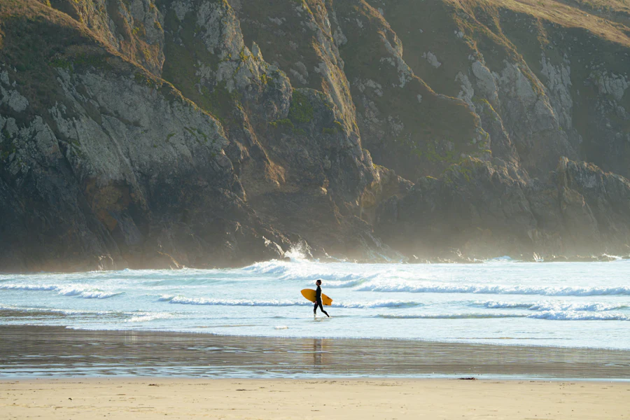 Surfer walking along the shoreline with a surfboard, framed by rugged cliffs and gentle waves. The scene captures a tranquil moment in a coastal paradise.