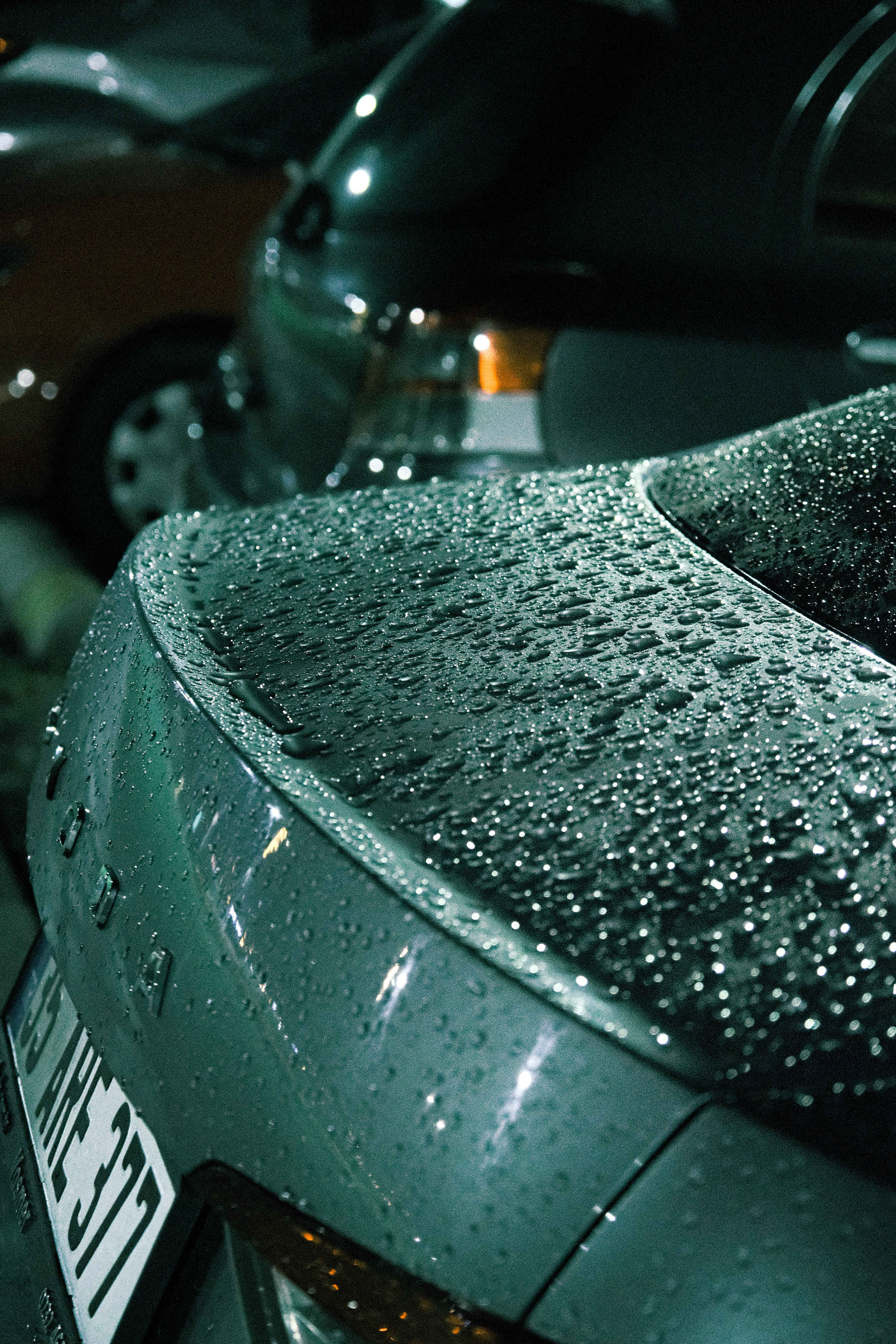 Close-up of a car’s rear with raindrops covering the surface, illuminated by artificial night lights, creating a cinematic urban mood. | Raindrops on a car's trunk at night